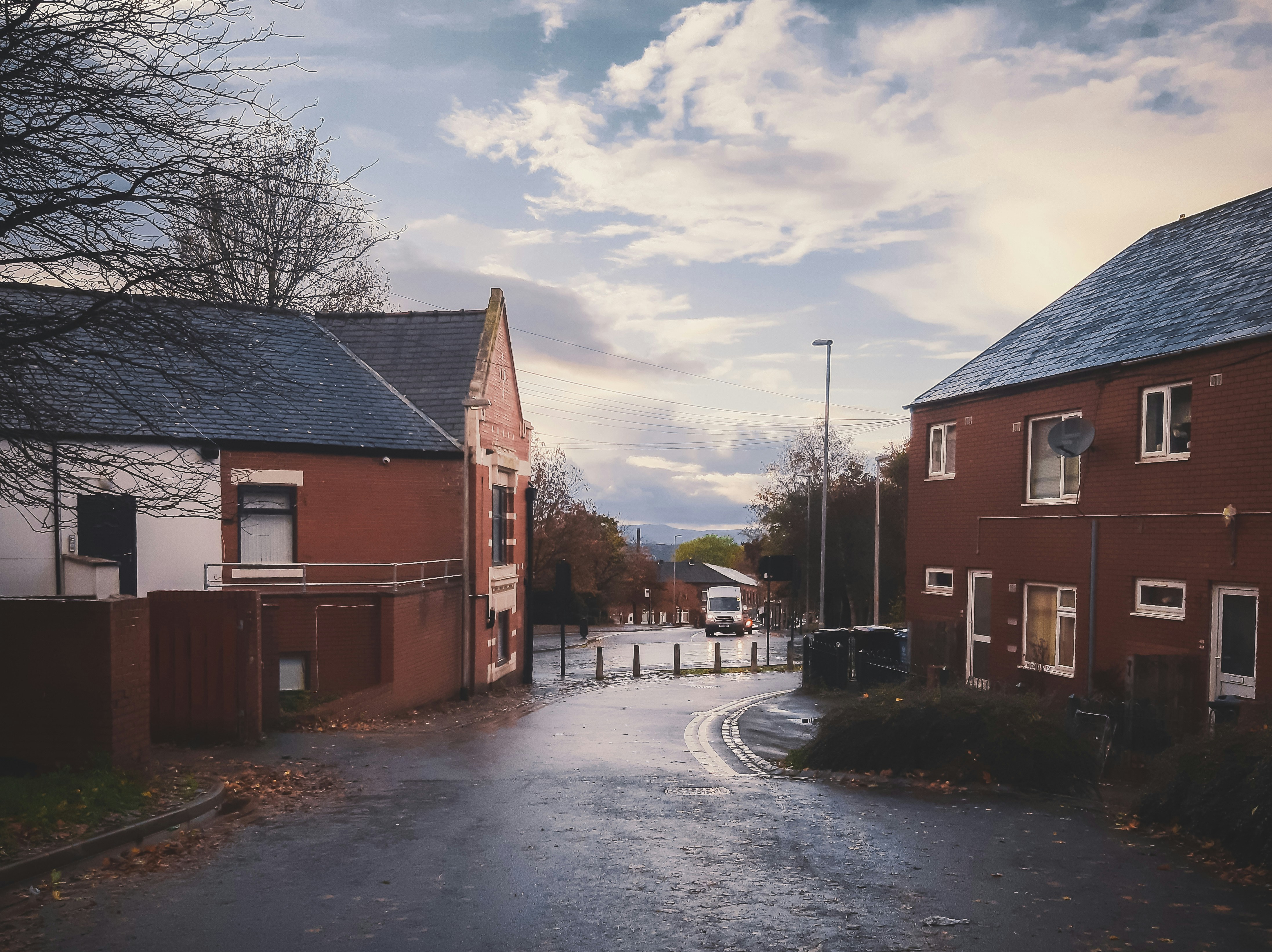 A tranquil street scene at twilight, featuring a gentle curve in the road bordered by residential buildings and autumn foliage. The sky is painted with soft hues of dusk.