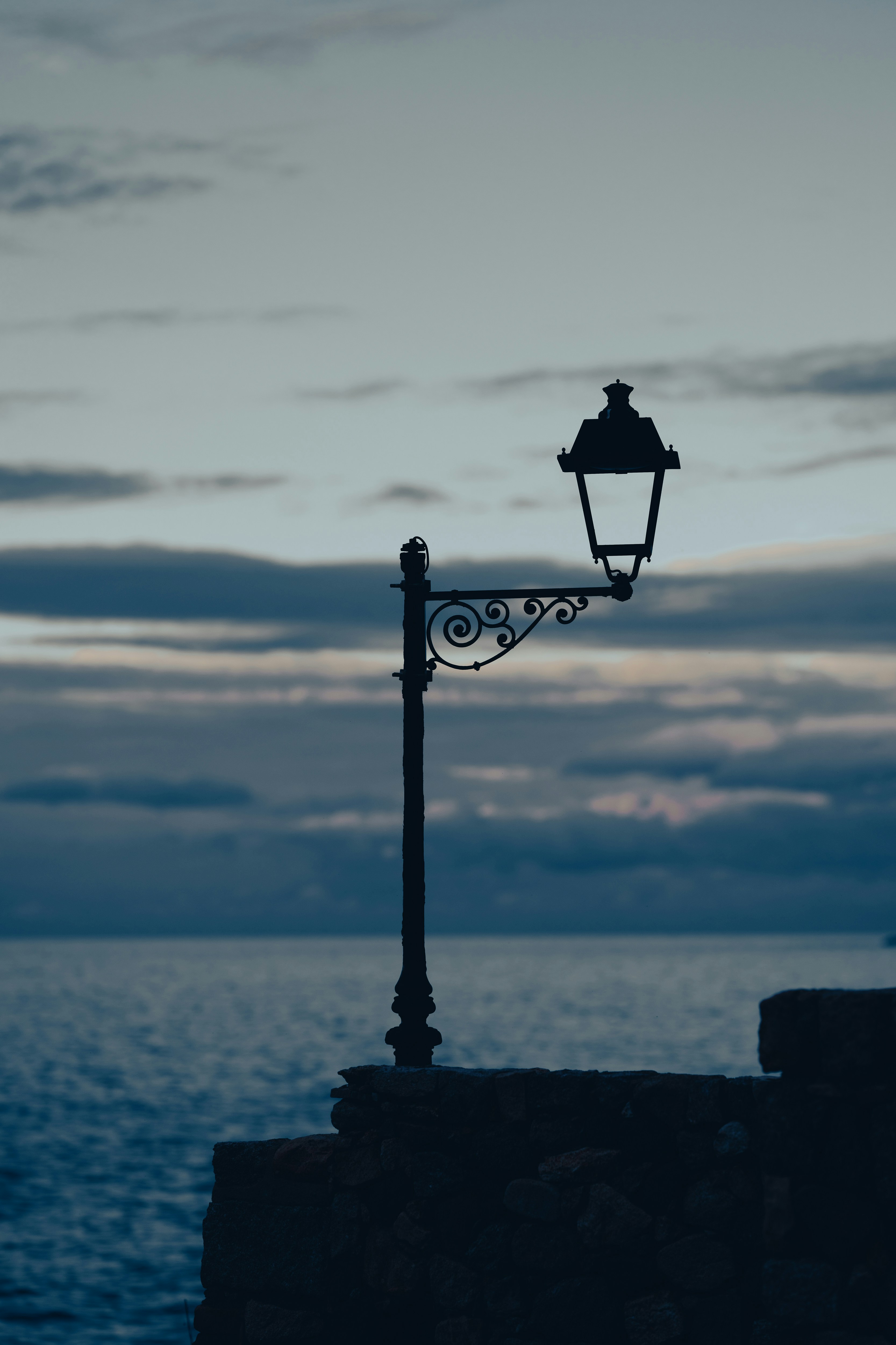 Silhouette of an ornate street lamp against a tranquil sea at dusk, capturing the serene transition from day to night.