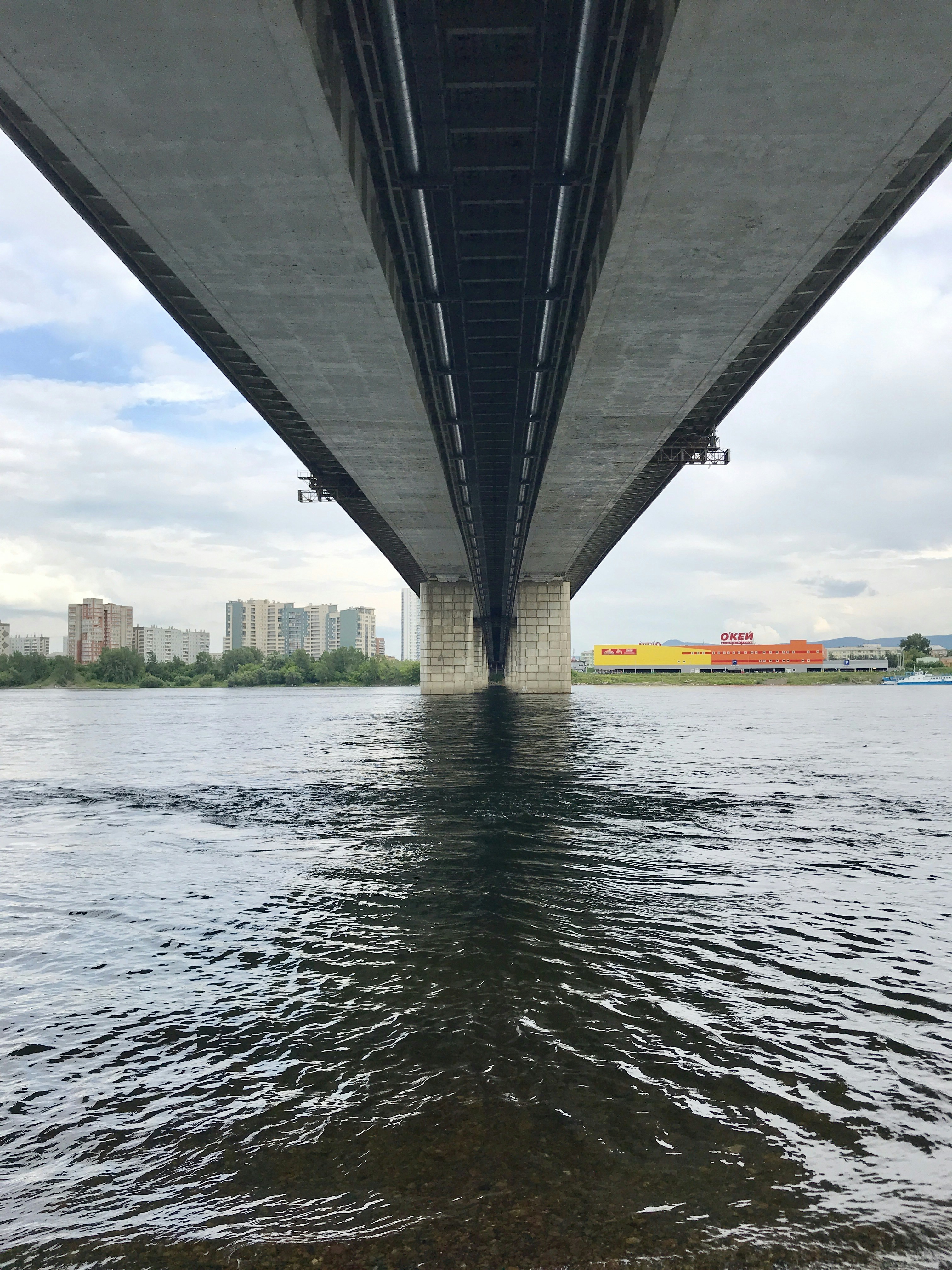 Concrete bridge viewed from underneath, reflecting in the water below with urban structures in the background.