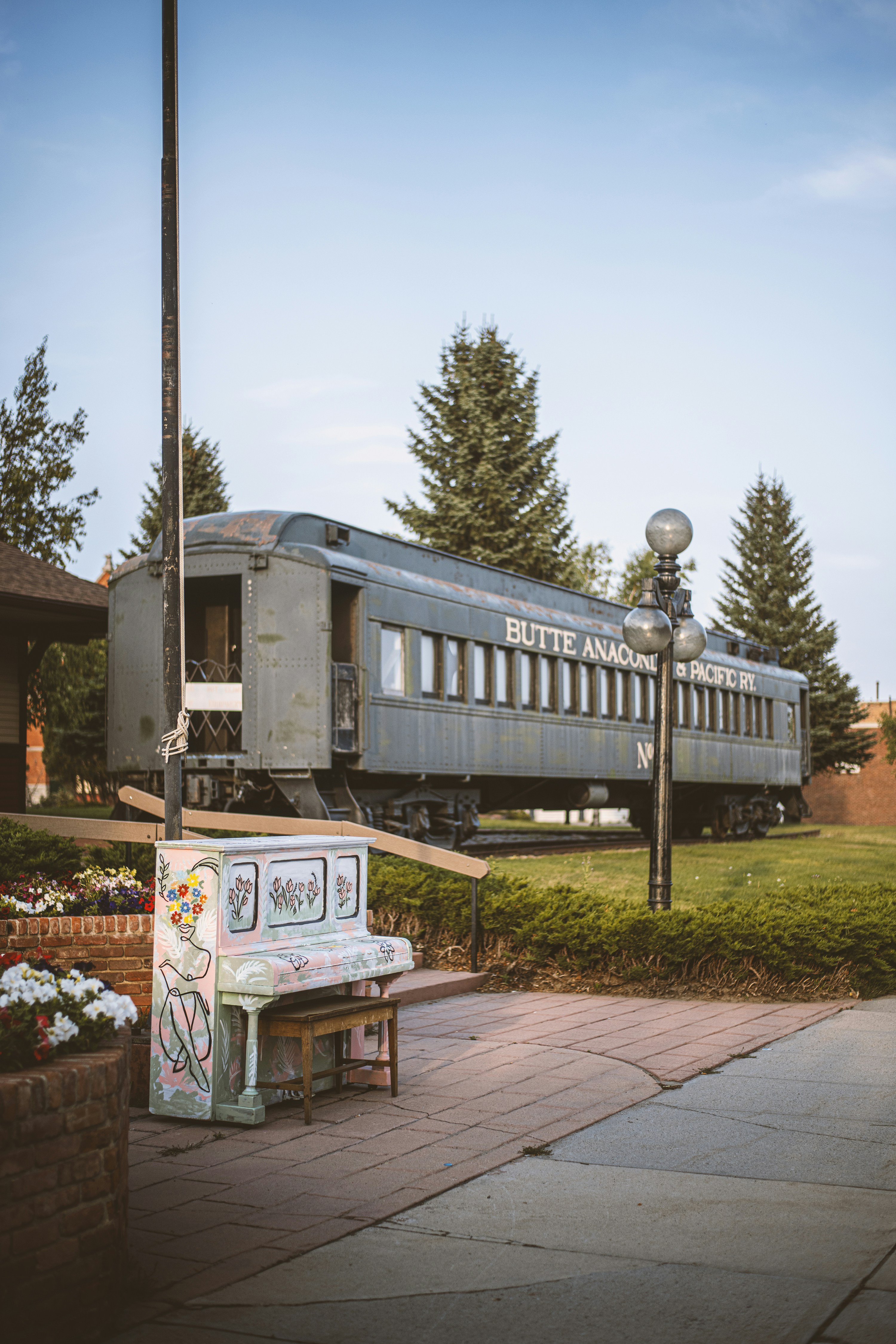 A historic train car stands prominently in a park, surrounded by greenery and vibrant flowers, with a whimsical painted piano nearby.
