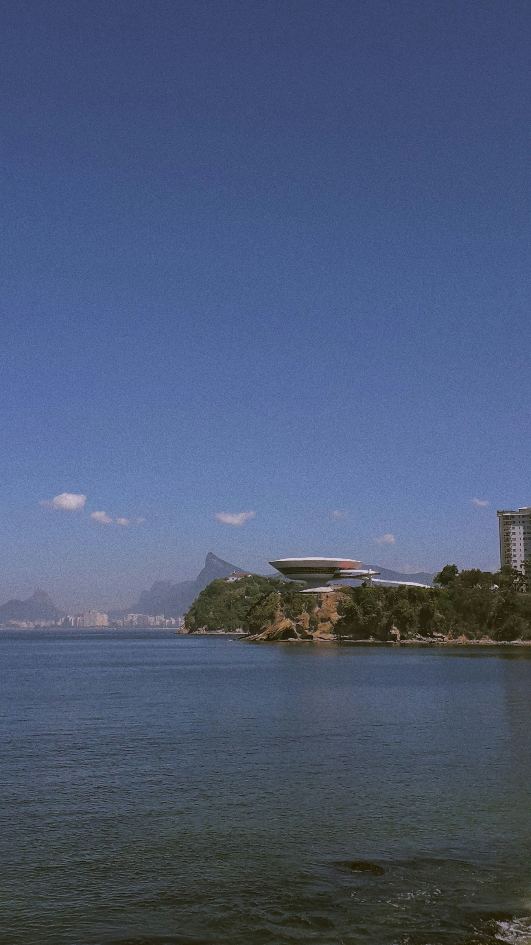 A photograph of a calm coastal scene featuring a rounded disc-shaped pavilion perched on a hillside overlooking a tranquil bay beneath a clear blue sky.