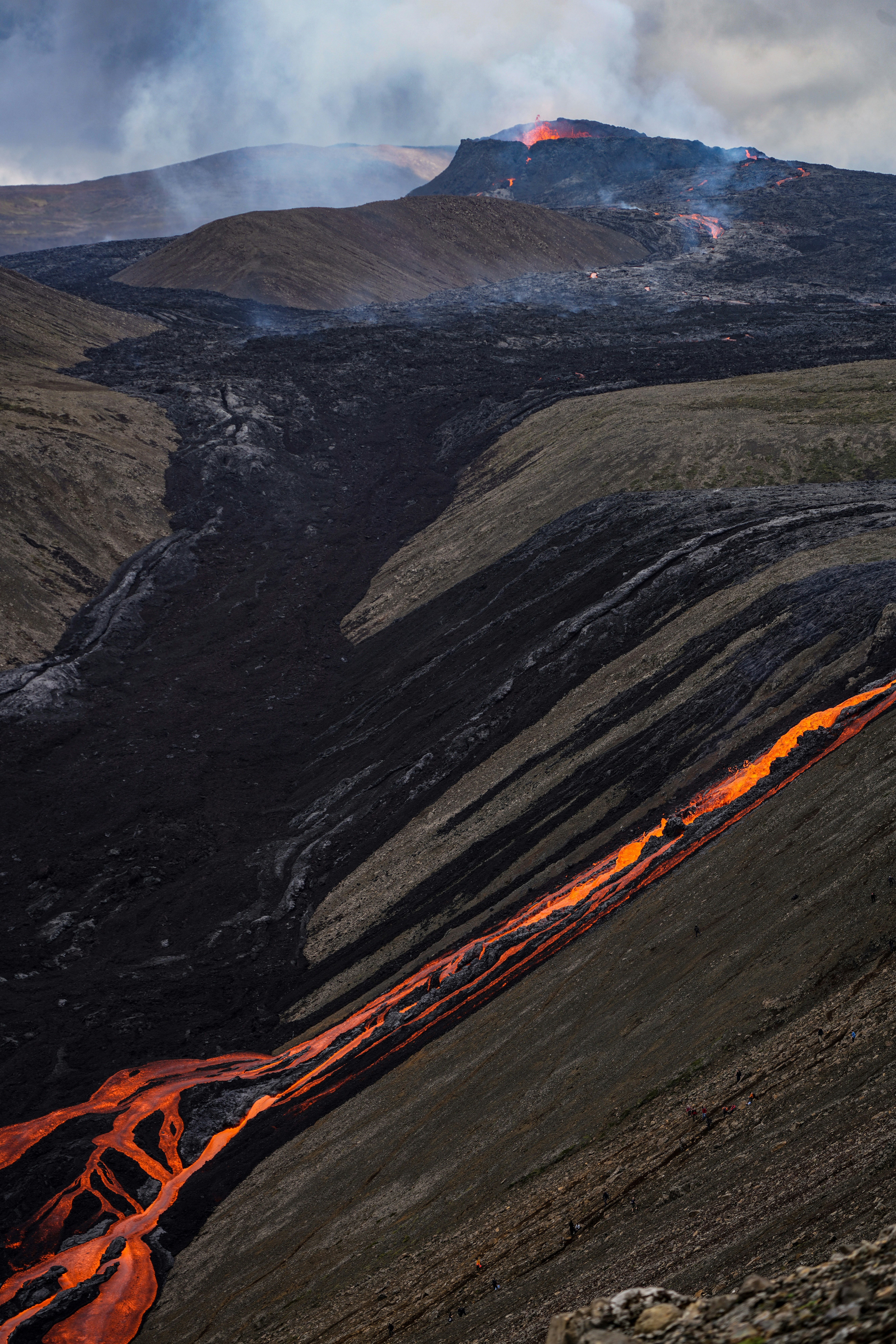 Lava flows gracefully down a rugged volcanic landscape, surrounded by dark, hardened rock and wisps of smoke rising from the summit. 
