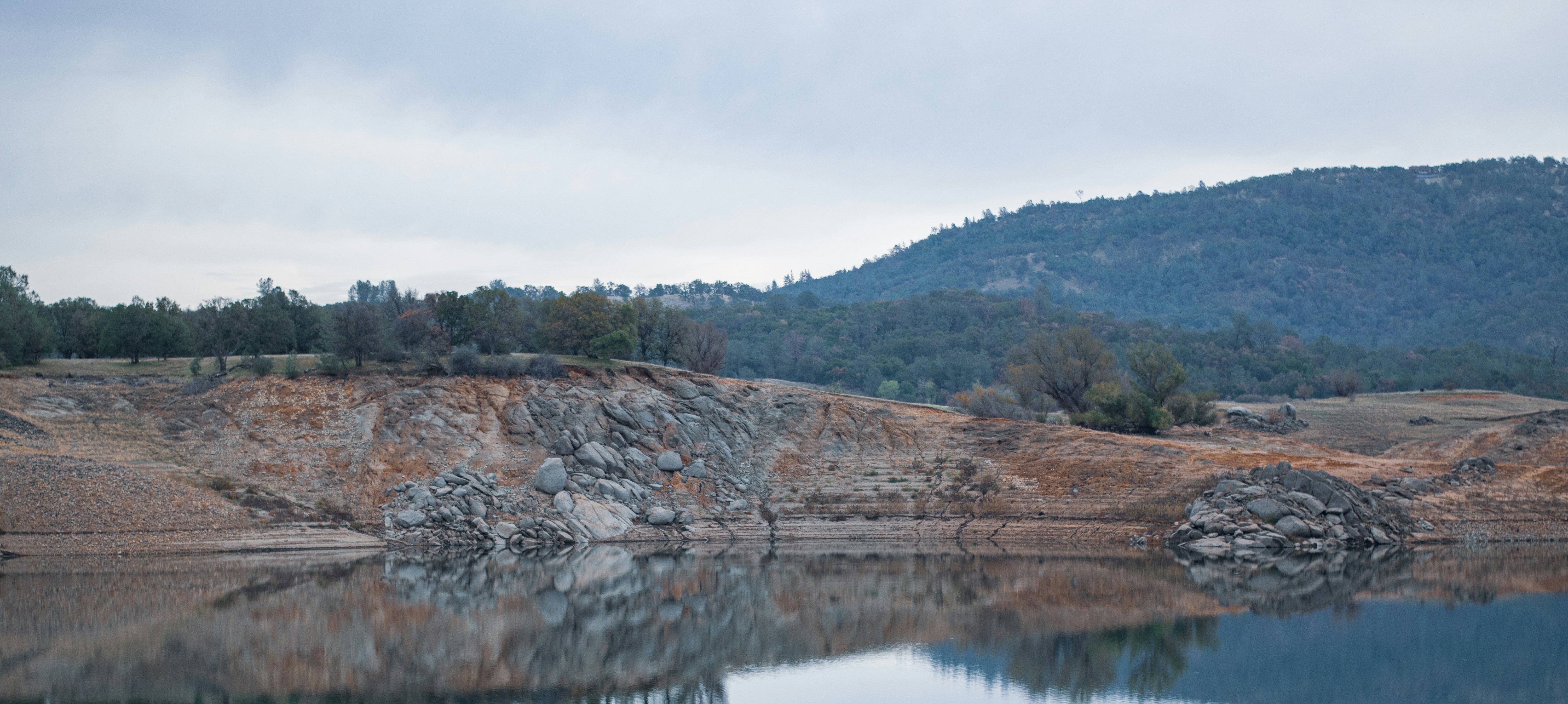 Tranquil body of water with rugged mountains and trees reflected under a cloudy sky.