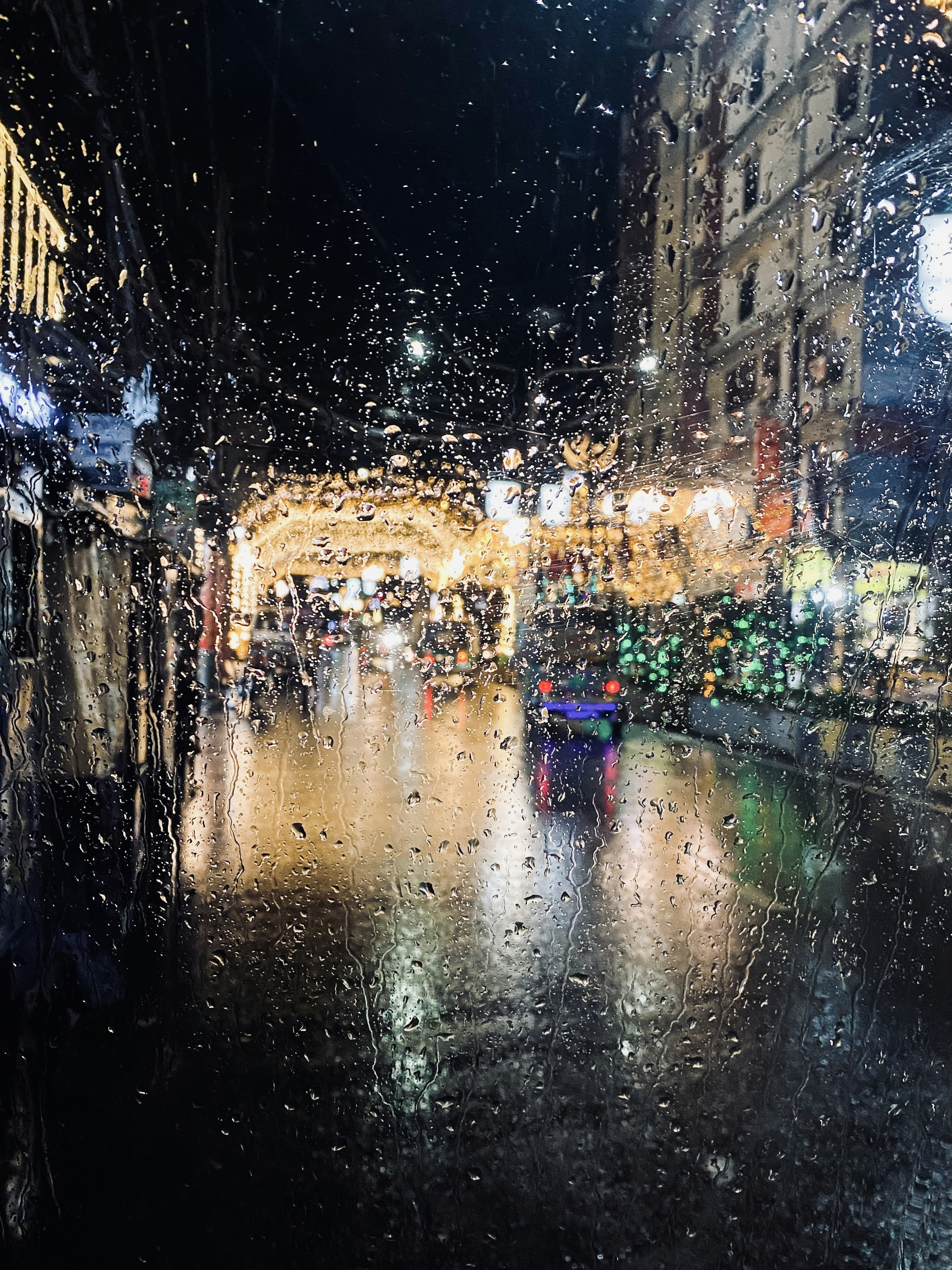 Raindrops cling to a window, blurring the vibrant lights of a bustling street decorated for the night. The scene captures the essence of urban life in the rain.