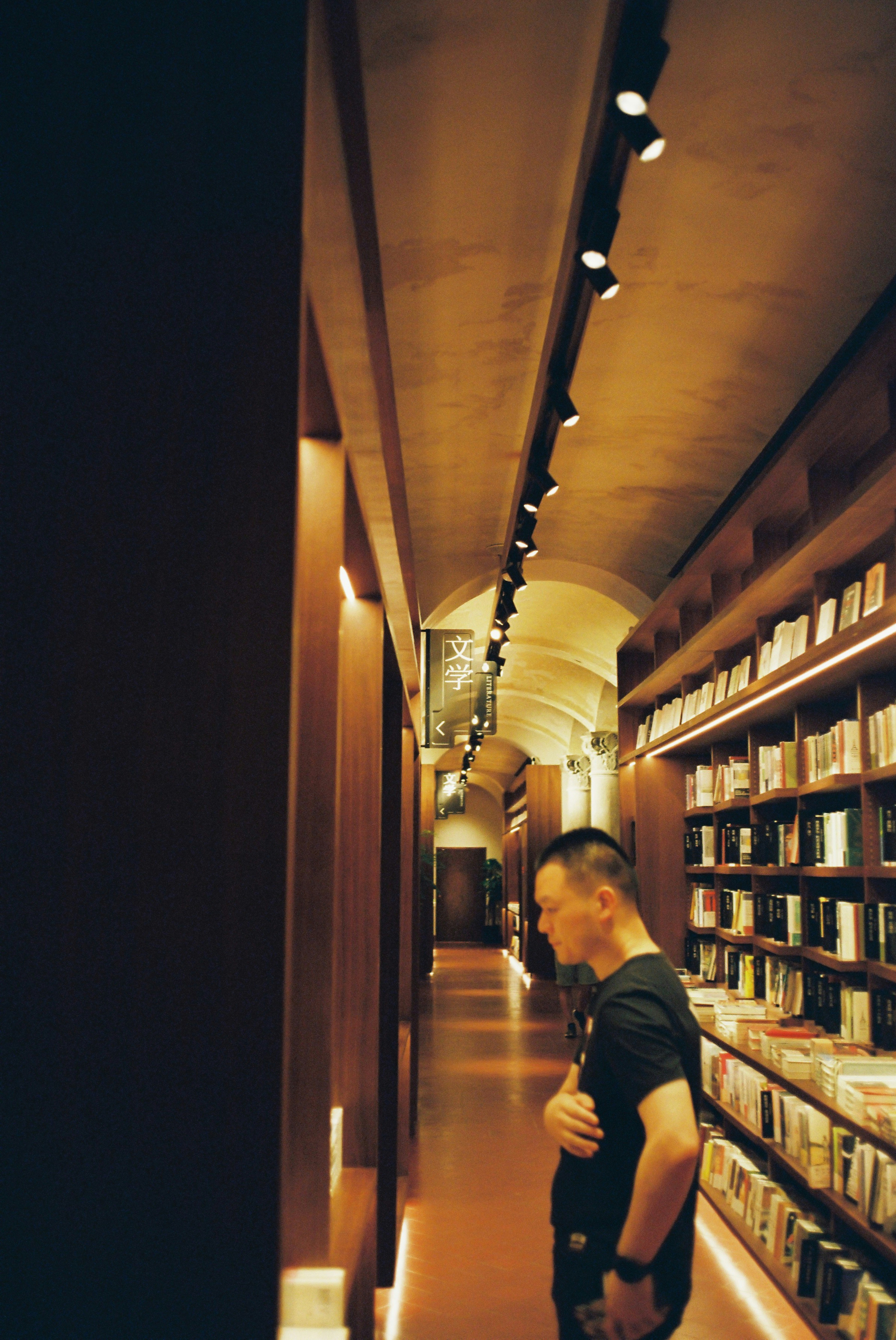 A contemplative figure stands in a warmly lit library corridor lined with bookshelves, creating an inviting atmosphere for exploration.