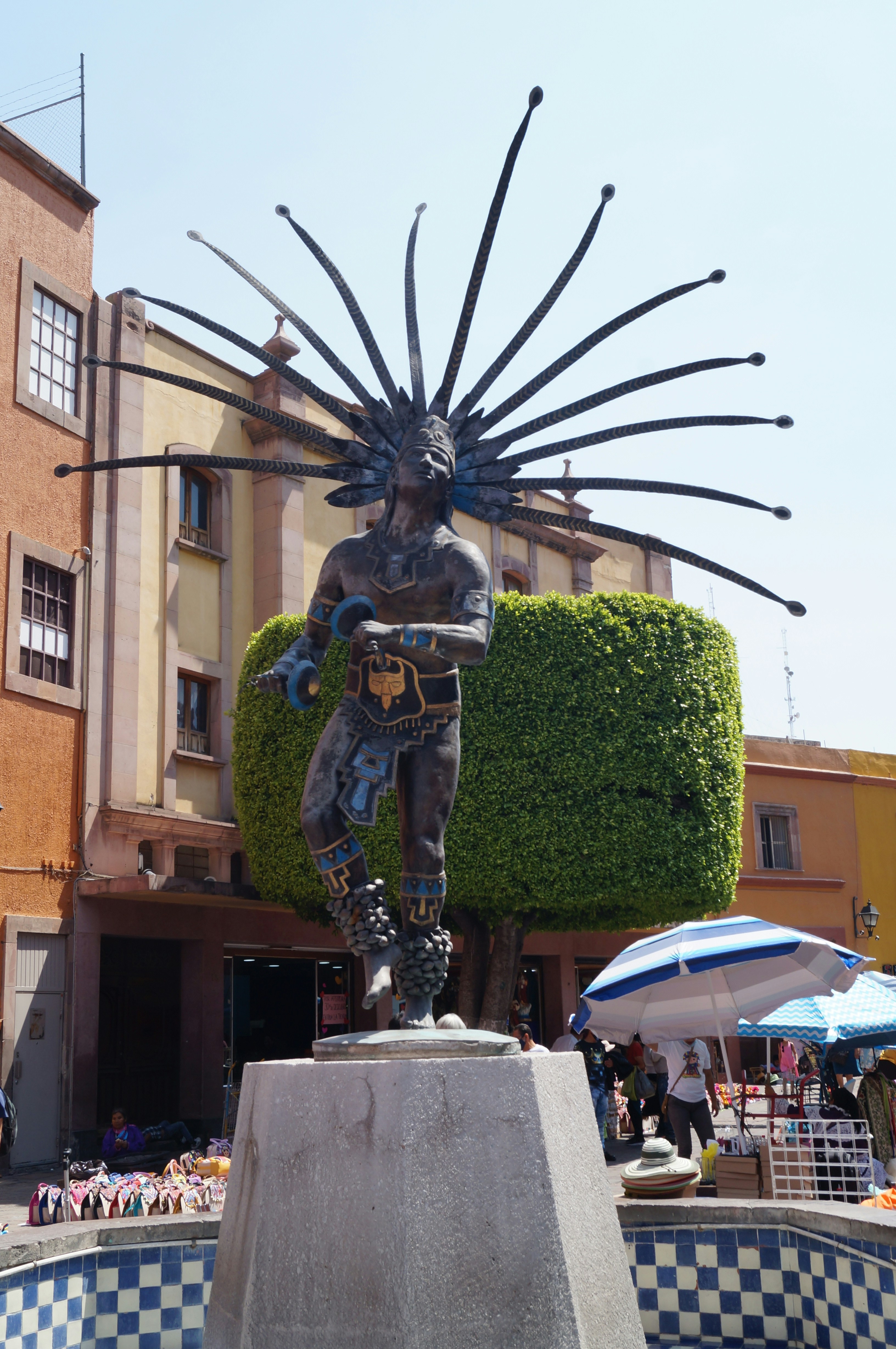 Bronze statue of a warrior adorned with traditional attire, surrounded by vibrant urban life and greenery. The sculpture stands proudly in a lively market square.
