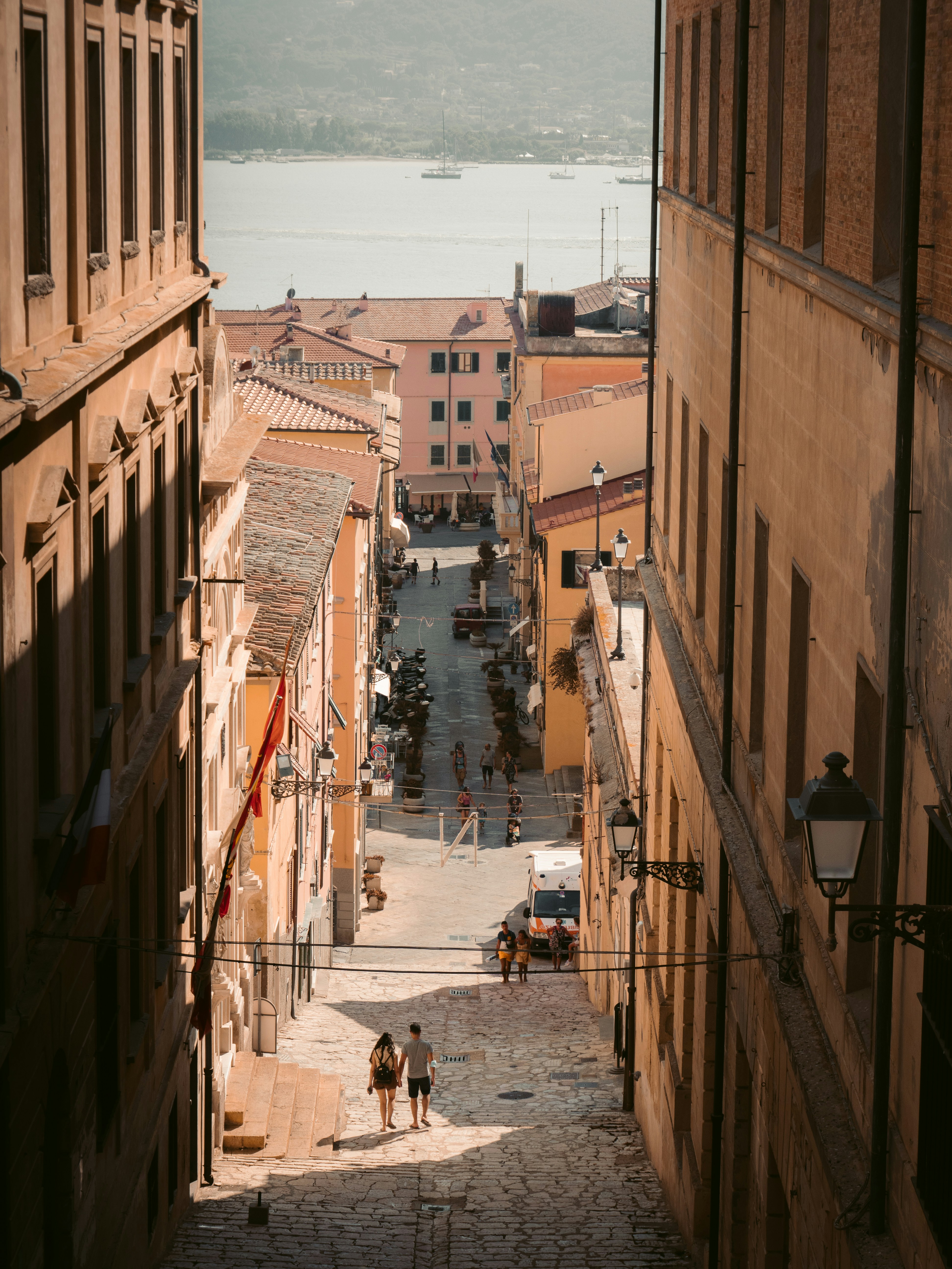 Sunlit alley descends between warm ochre façades toward a harbor, with pedestrians visible along the cobbled steps.