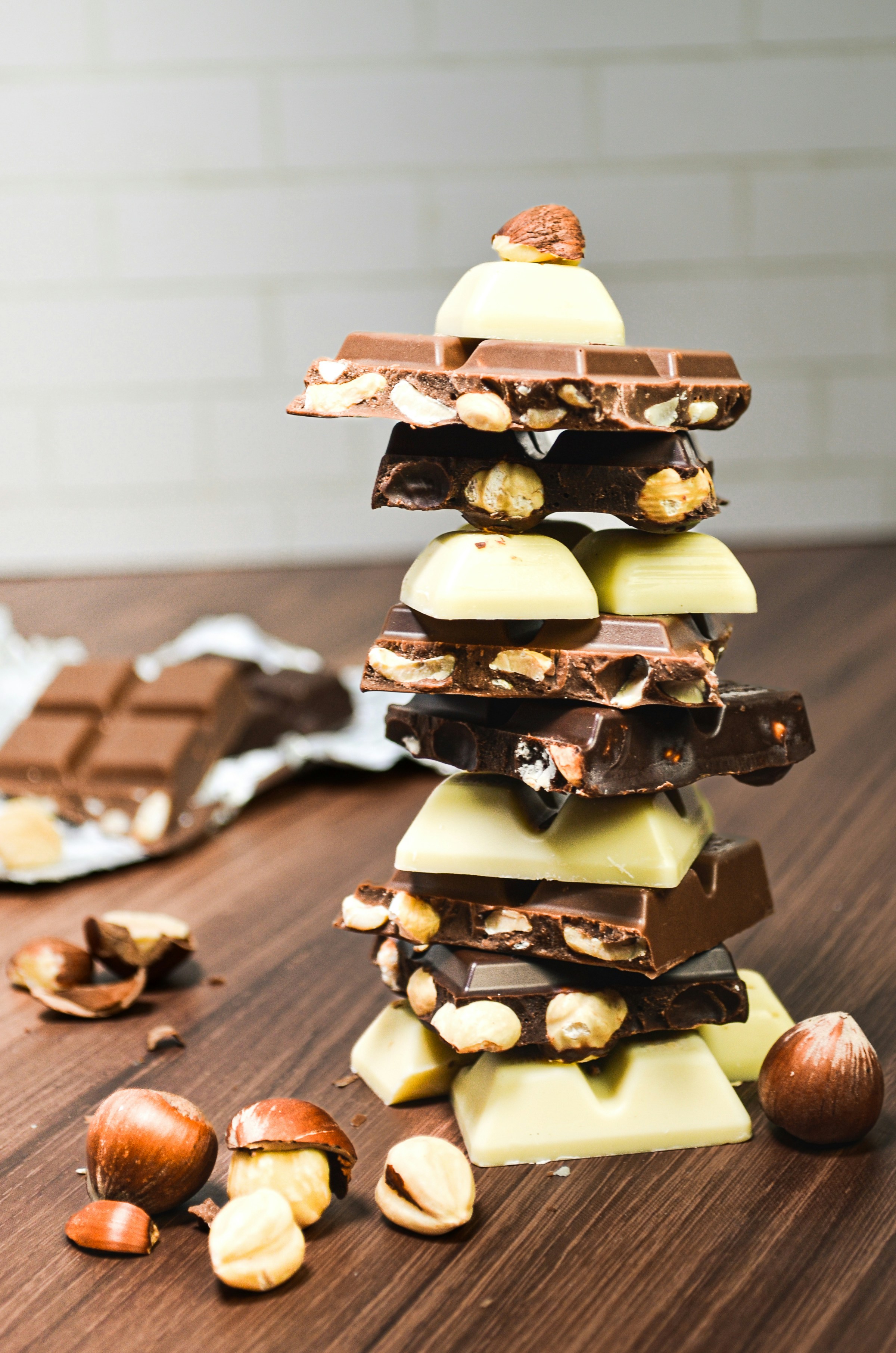 A carefully arranged stack of assorted chocolate bars topped with a hazelnut, surrounded by scattered nuts. The warm wooden surface complements the rich textures of the chocolates.