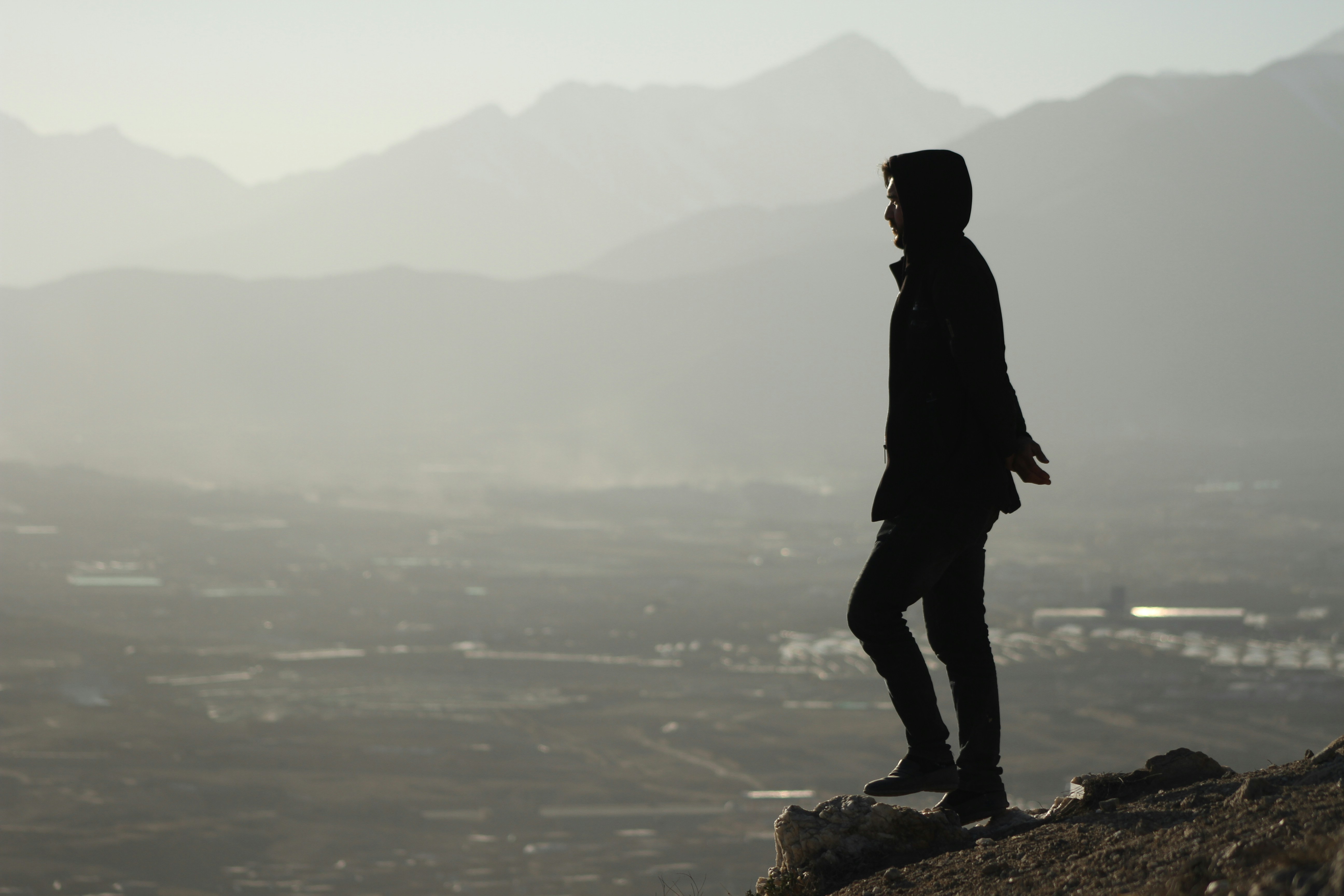 A solitary figure stands on a rocky outcrop, silhouetted against a hazy landscape of mountains and valleys. The scene conveys a sense of contemplation and connection with nature.