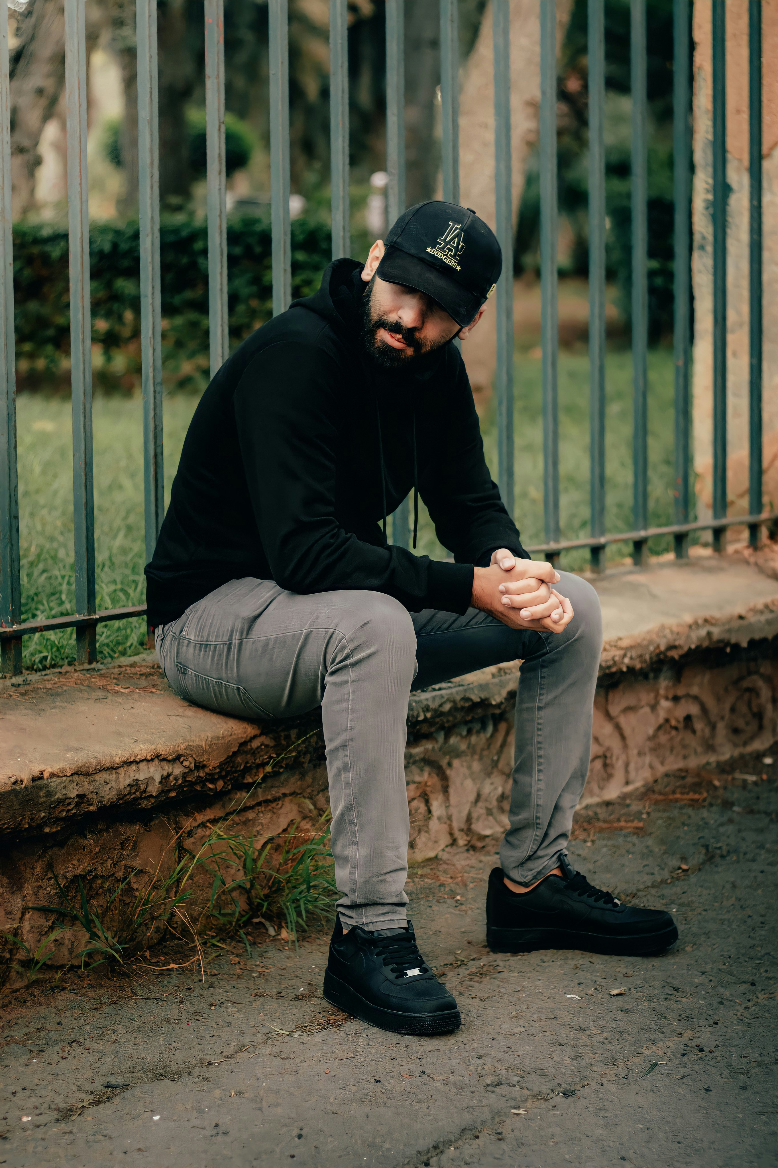 Man in a black hoodie and cap sitting pensively on a stone ledge beside a green lawn and metal fence.