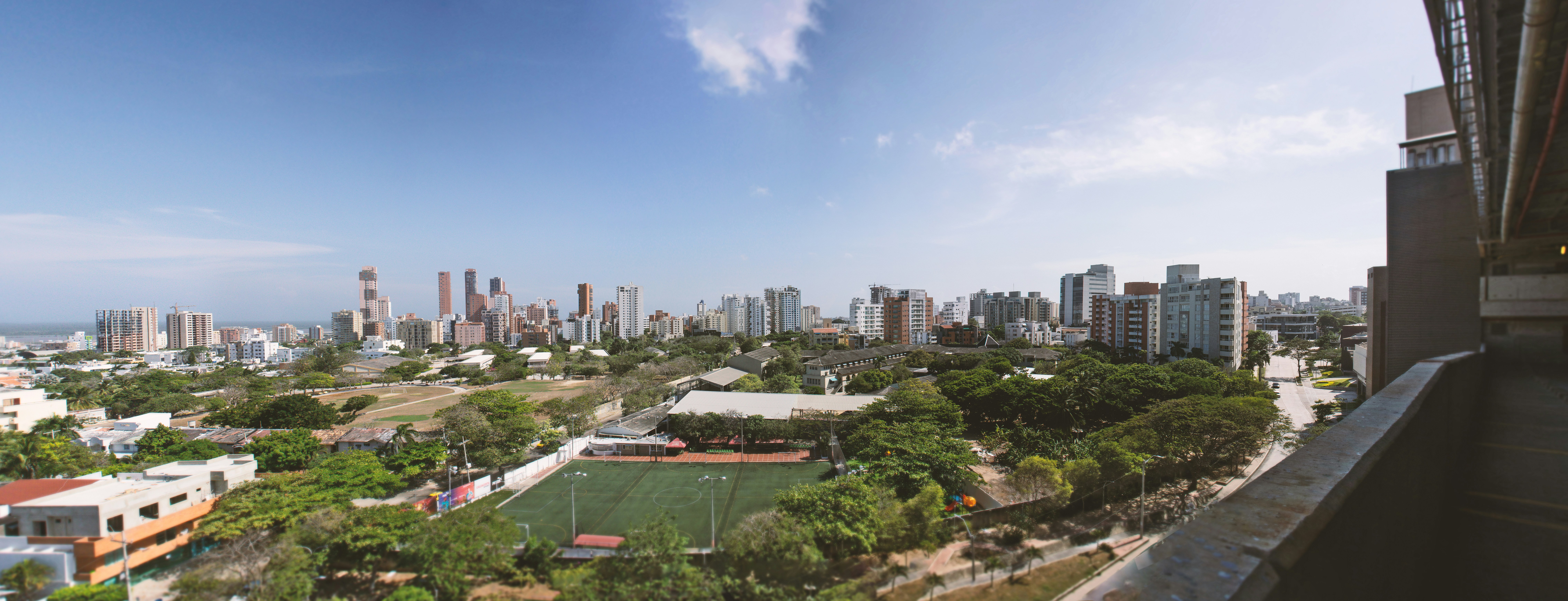 Panoramic view showcasing a city skyline with a lush soccer field in the foreground under a bright blue sky.