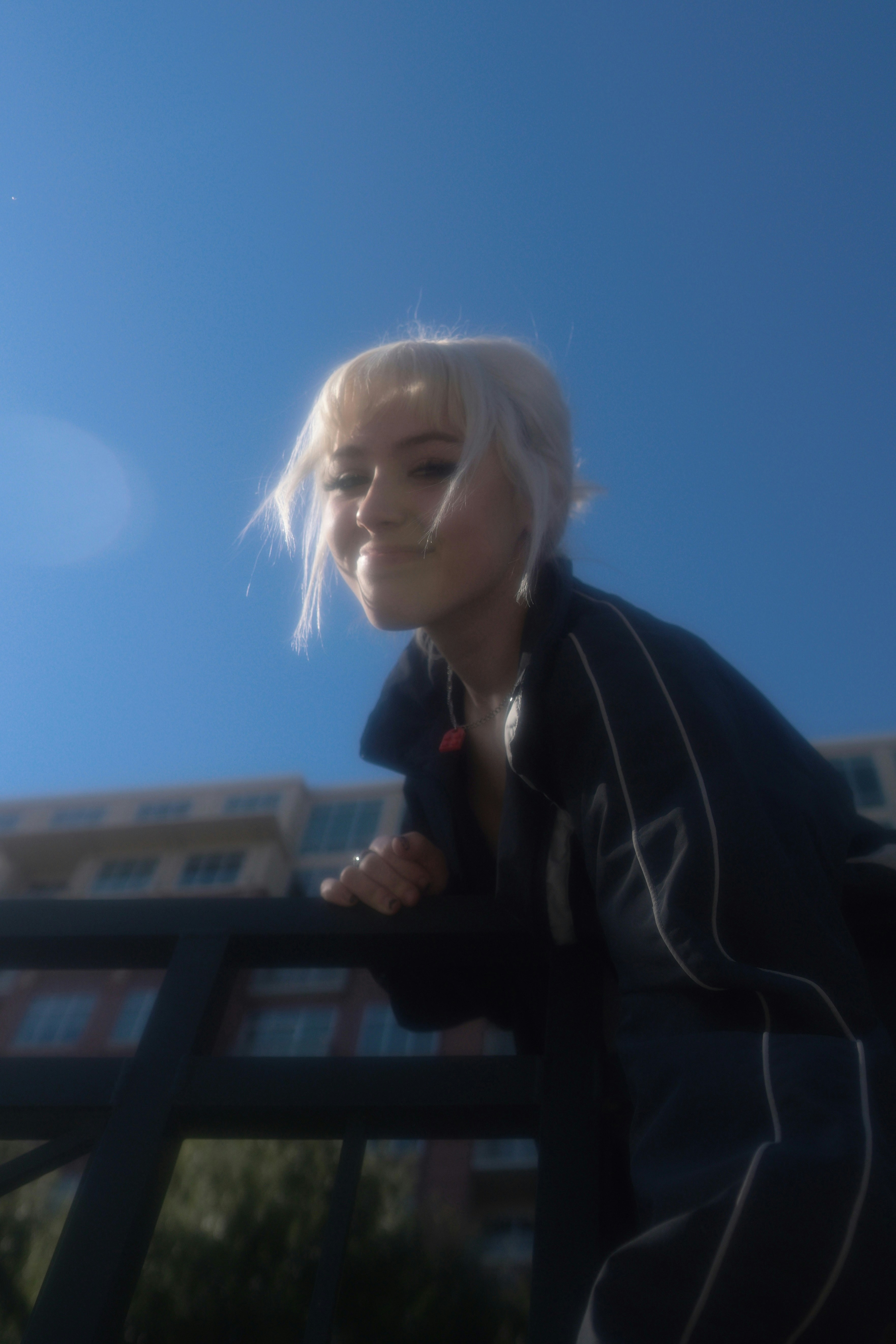 A young woman with short blonde hair leans over a railing, smiling against a bright blue sky. The urban backdrop adds depth to the cheerful moment.
