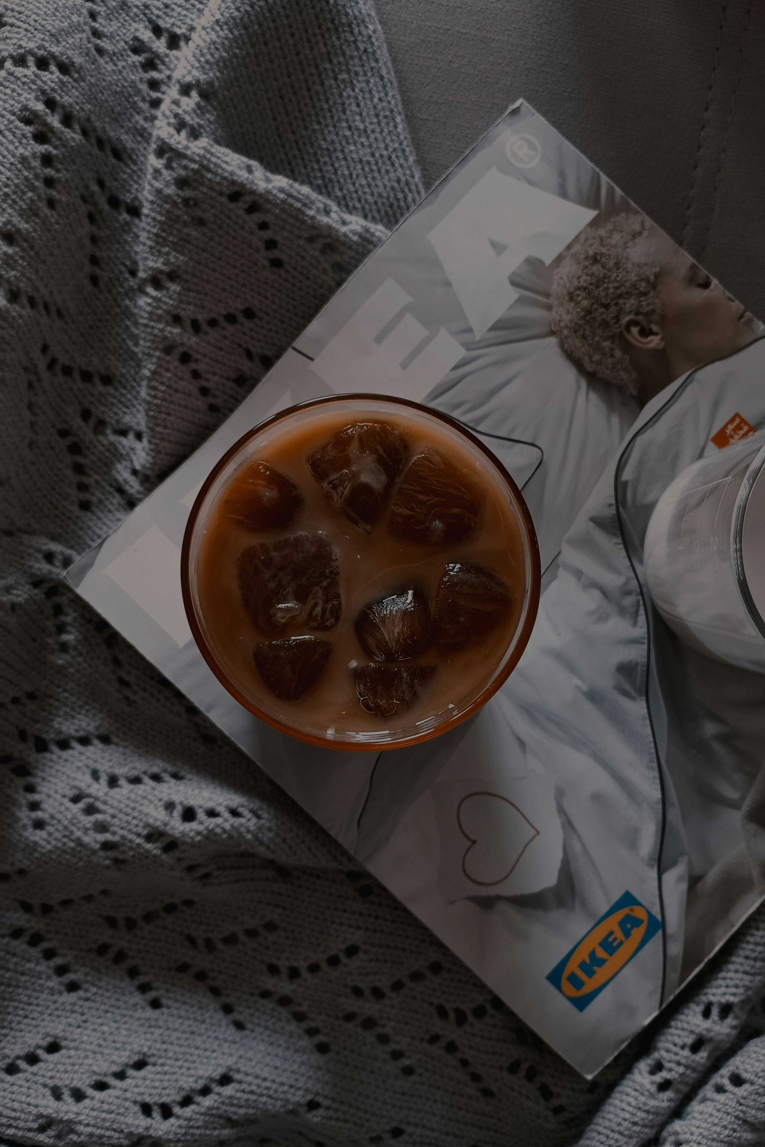 Iced coffee with ice cubes rests on a magazine, surrounded by a textured gray blanket.