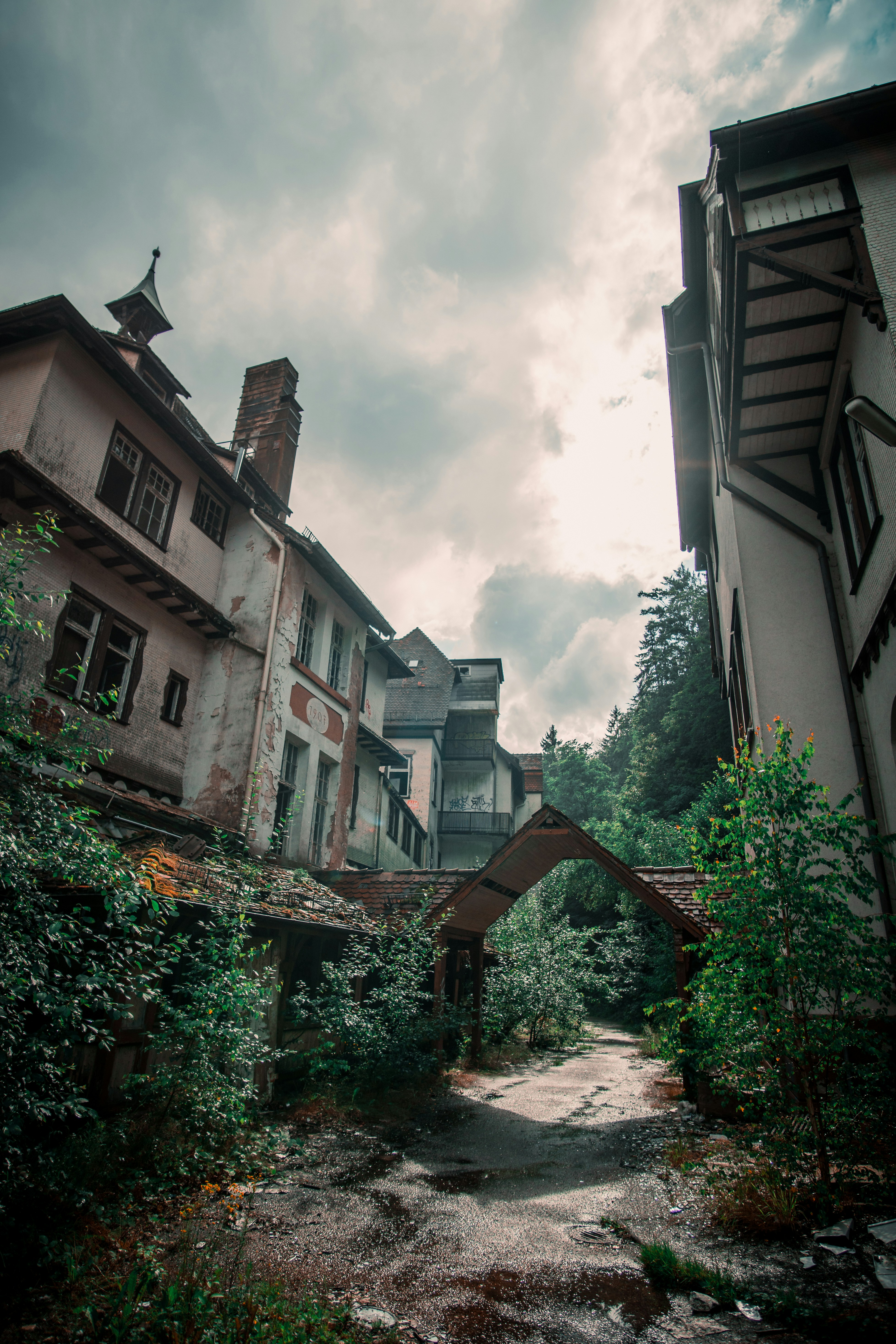Abandoned courtyard framed by decaying buildings, overgrown with vegetation and shrouded in moody clouds.