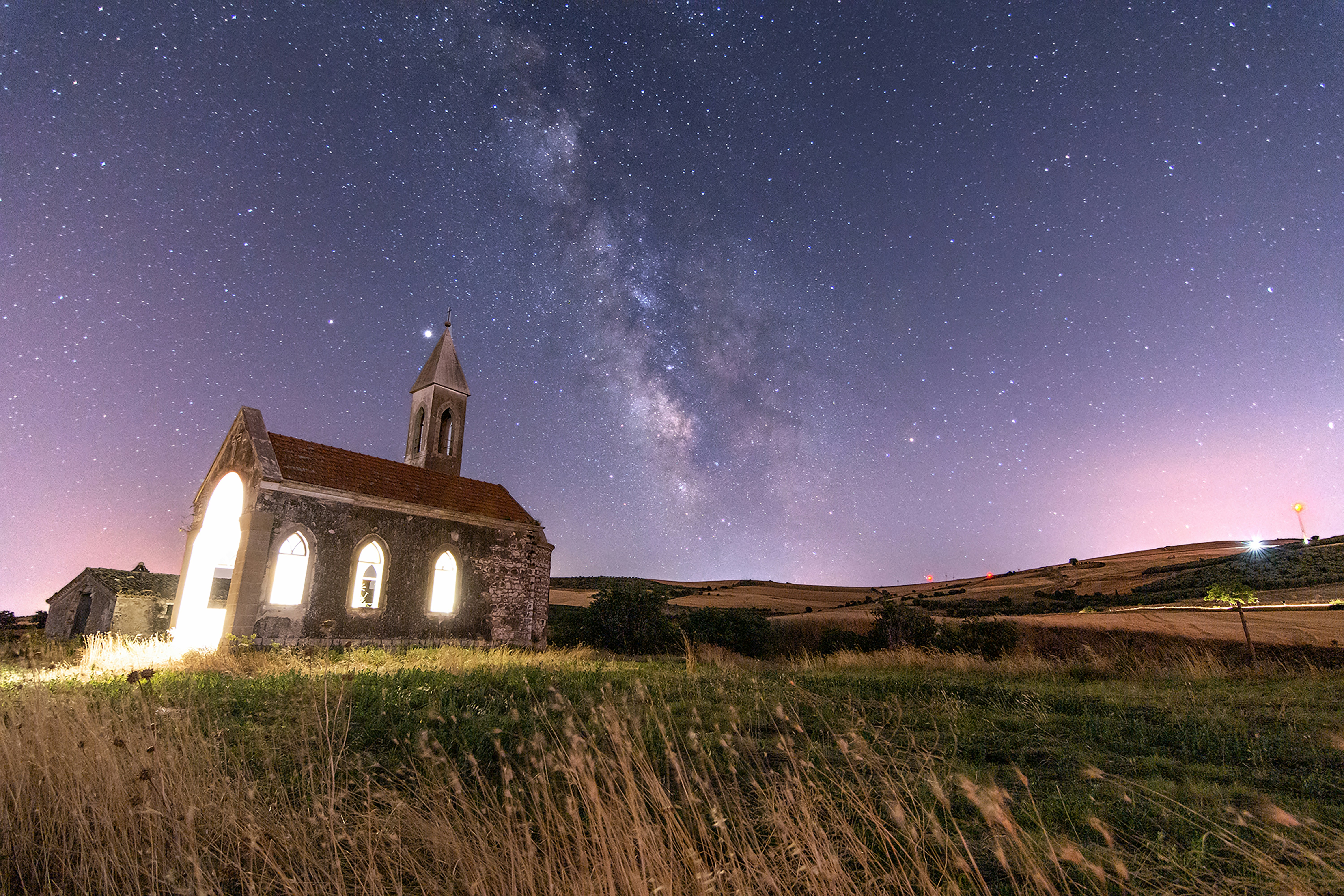 Illuminated chapel stands in a grassy field beneath a vibrant Milky Way sky.