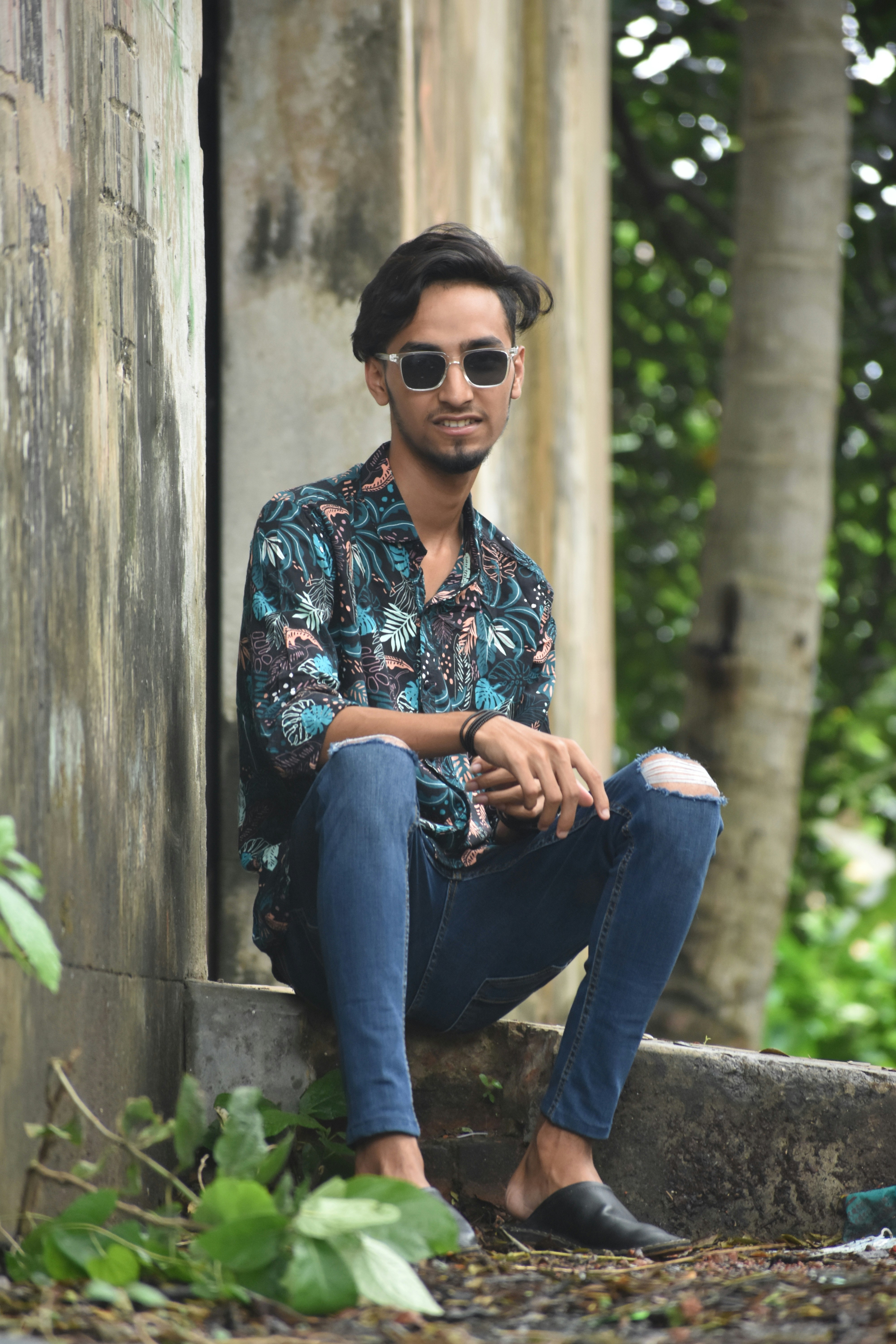 Young man in stylish attire sitting against a textured wall, surrounded by lush greenery. The scene conveys a relaxed, urban vibe.