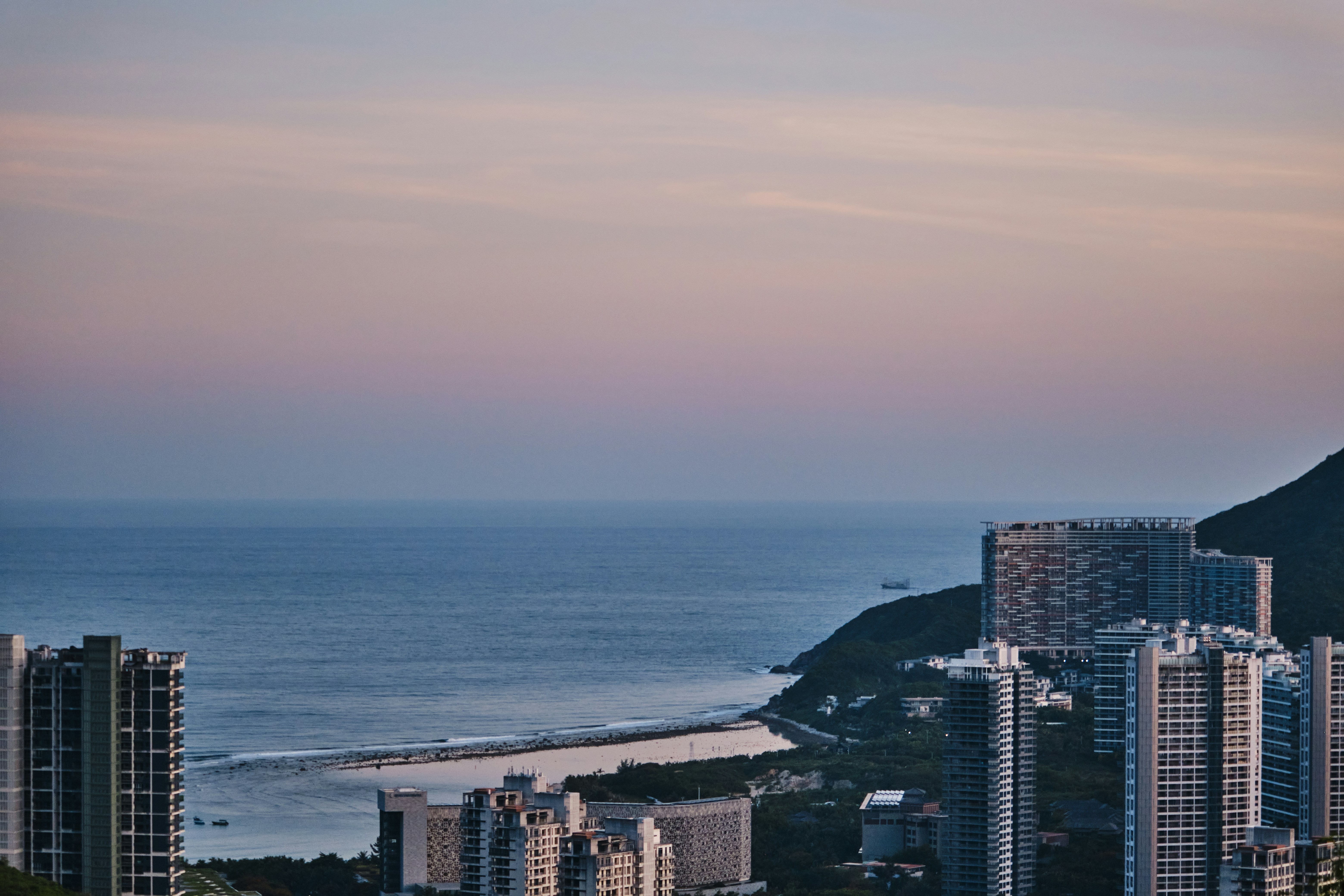 A panoramic view of a coastal city as the sun sets, highlighting modern skyscrapers against a tranquil sea and soft pastel sky.