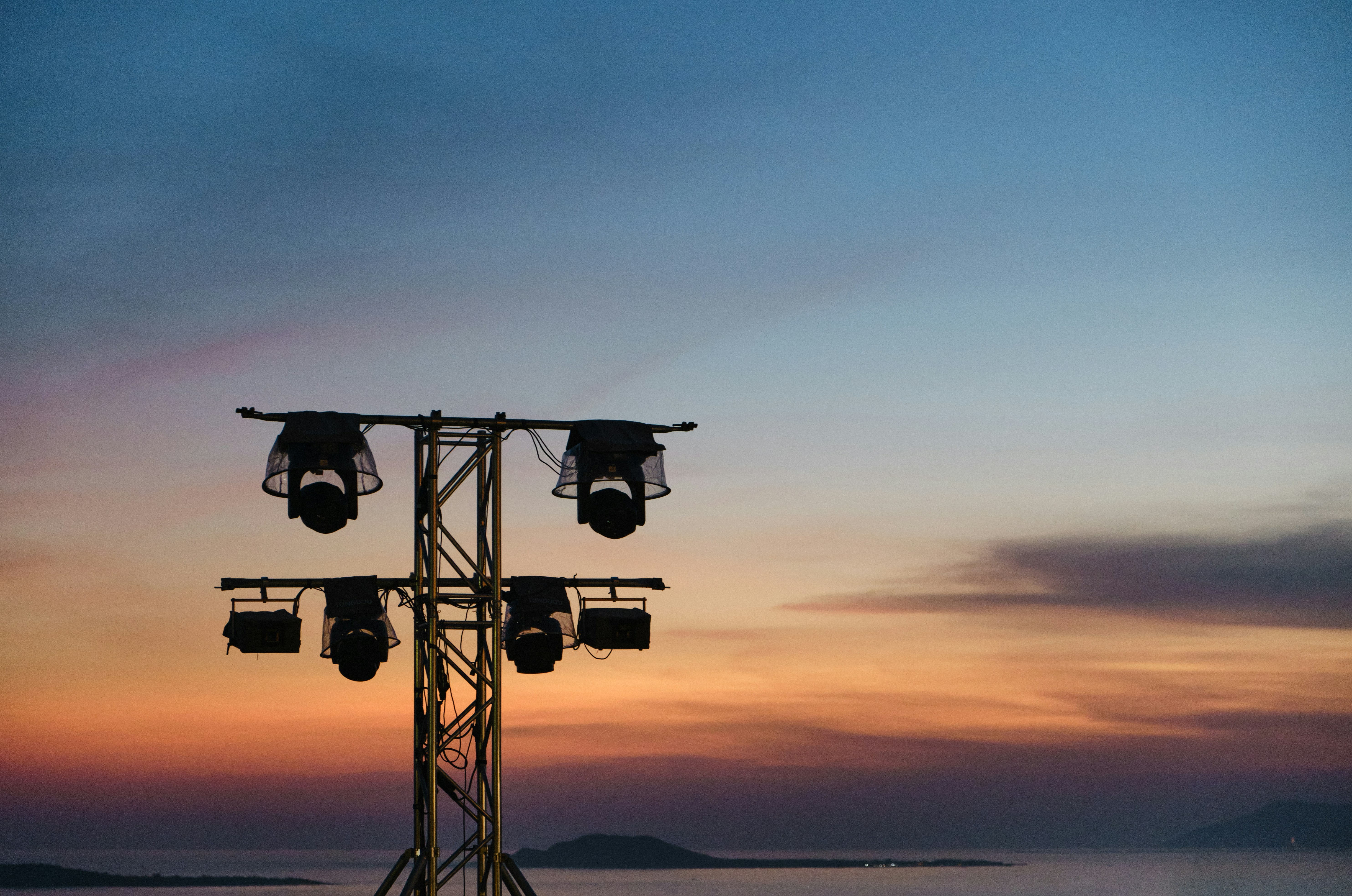 Stage lighting equipment silhouetted against a vibrant sunset sky, hinting at an upcoming event. The scene captures the anticipation of nightfall.