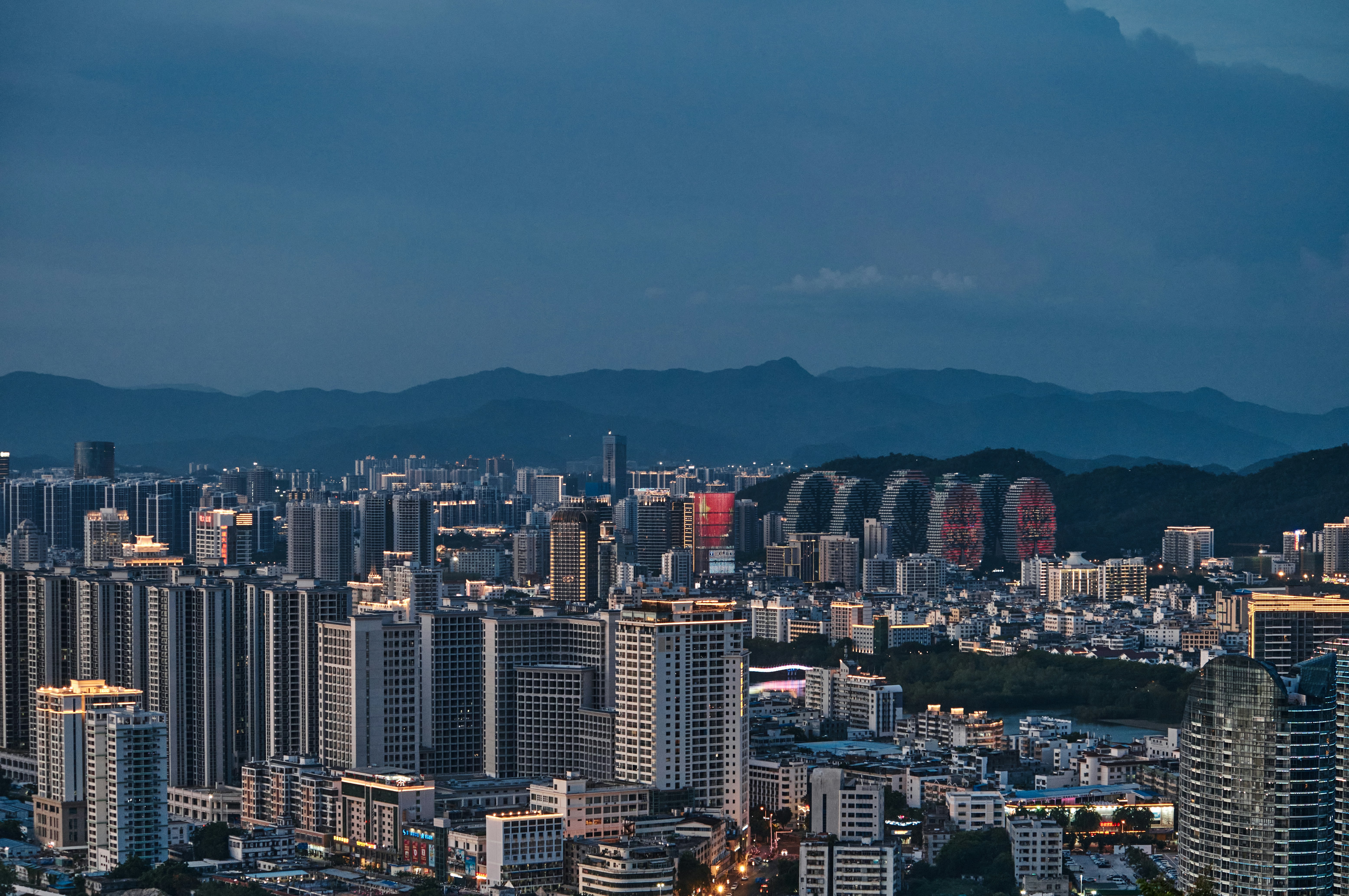 Vast cityscape illuminated under twilight, showcasing a blend of modern architecture and distant mountains. Bright lights contrast against the darkening sky.