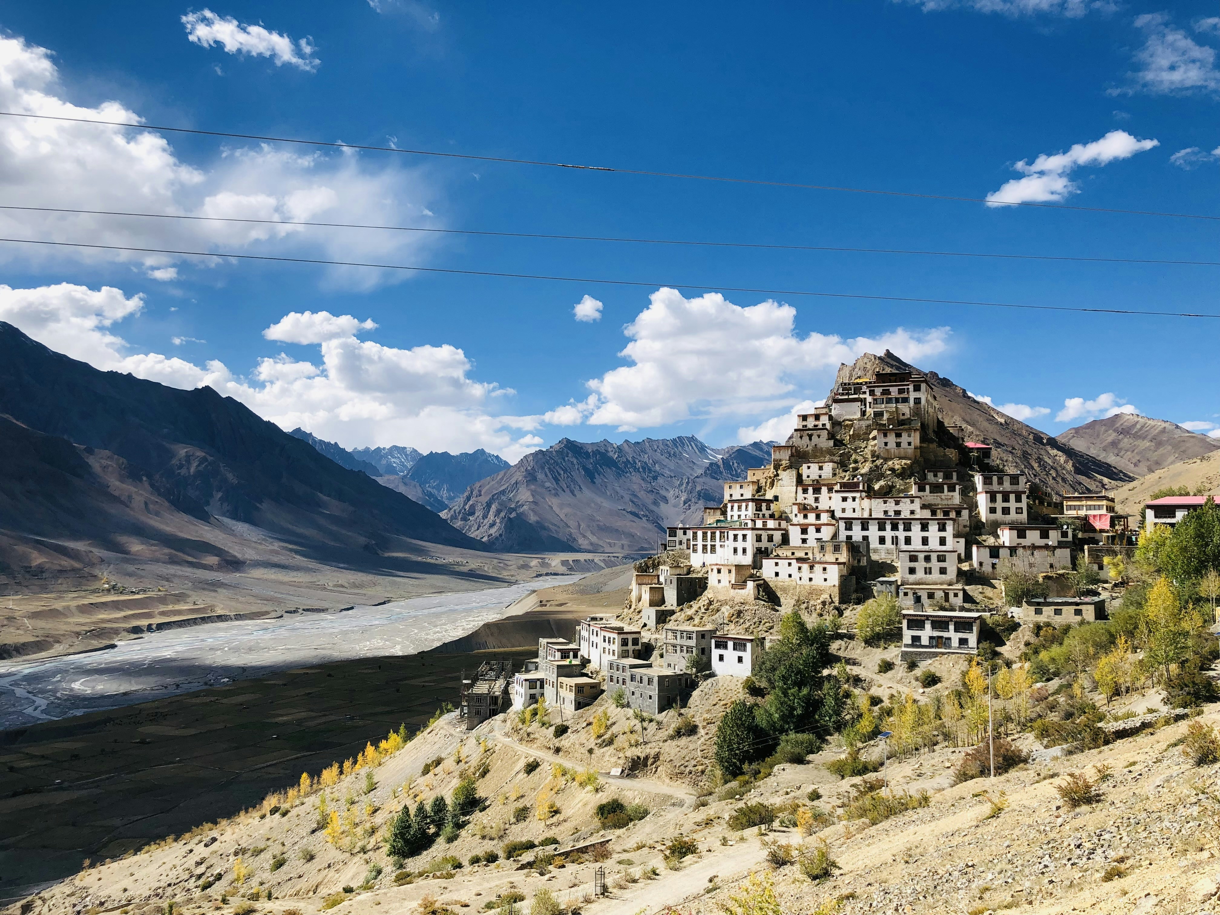 A historic monastery perched on a hillside overlooking a winding river and expansive valley, framed by towering mountains under a vibrant sky.