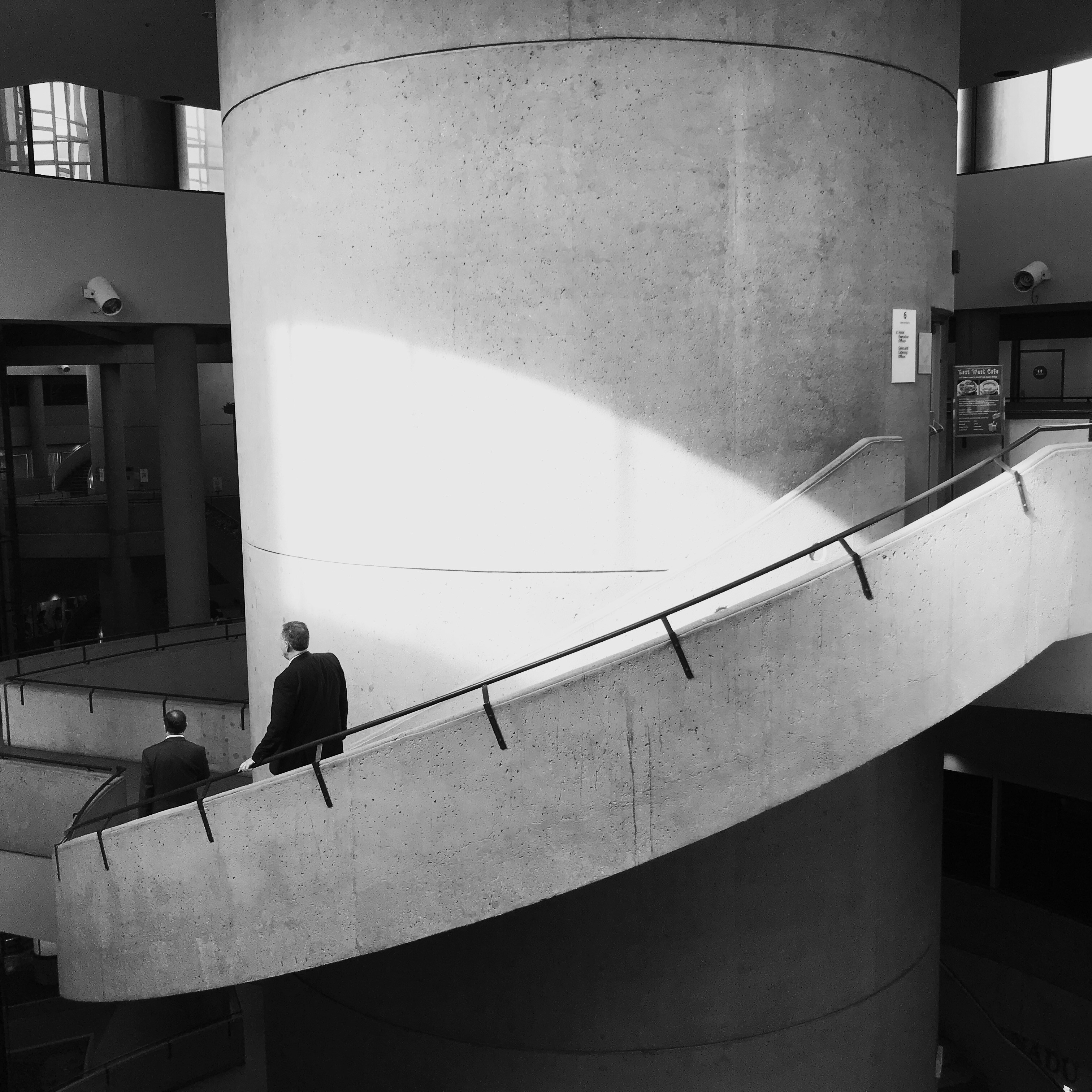 Two figures ascending a spiraling staircase within a modern architectural space, illuminated by a shaft of light. The stark contrast of black and white emphasizes the structural elements.