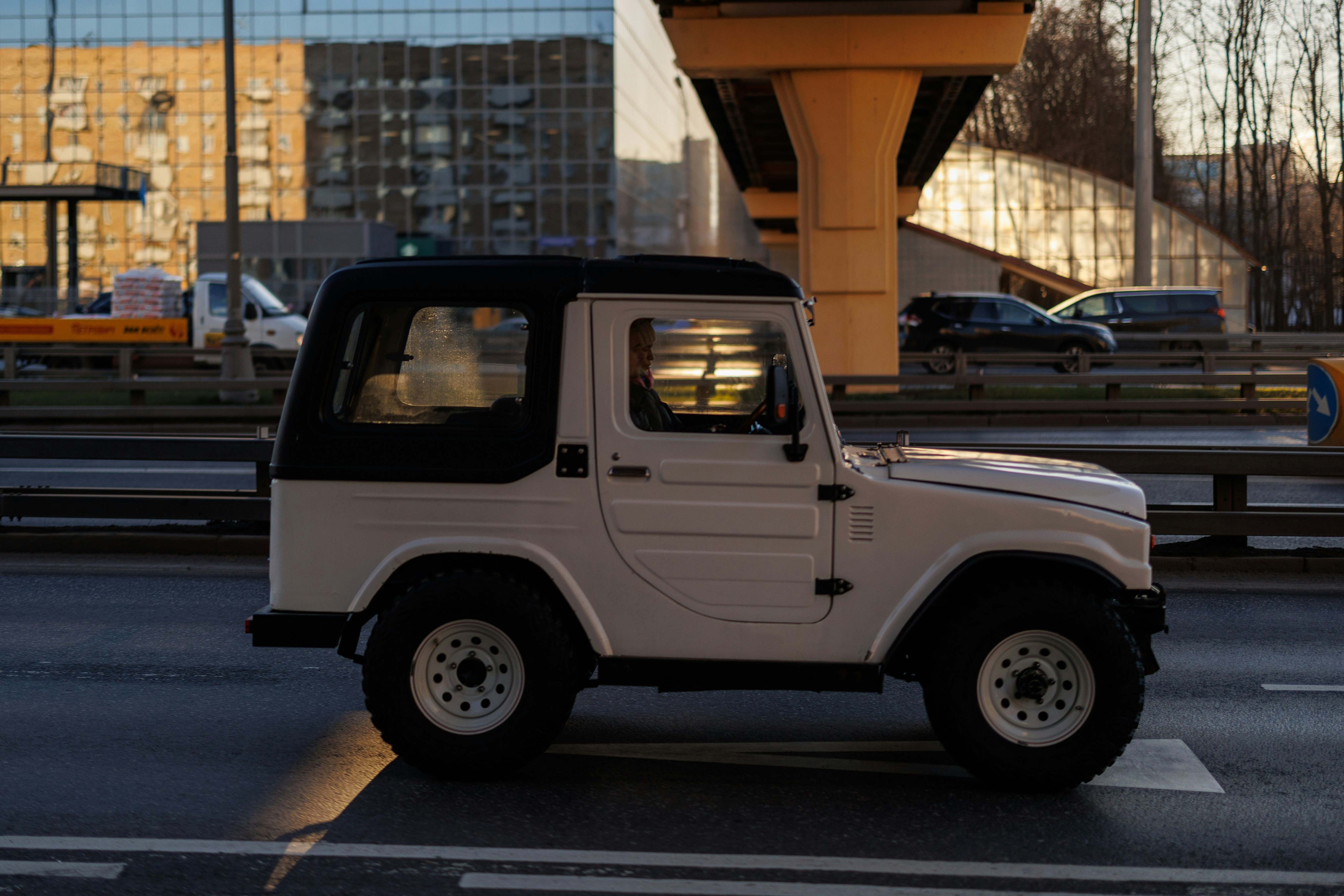 A white off-road vehicle traversing a city street, framed by modern architecture and late afternoon light.
