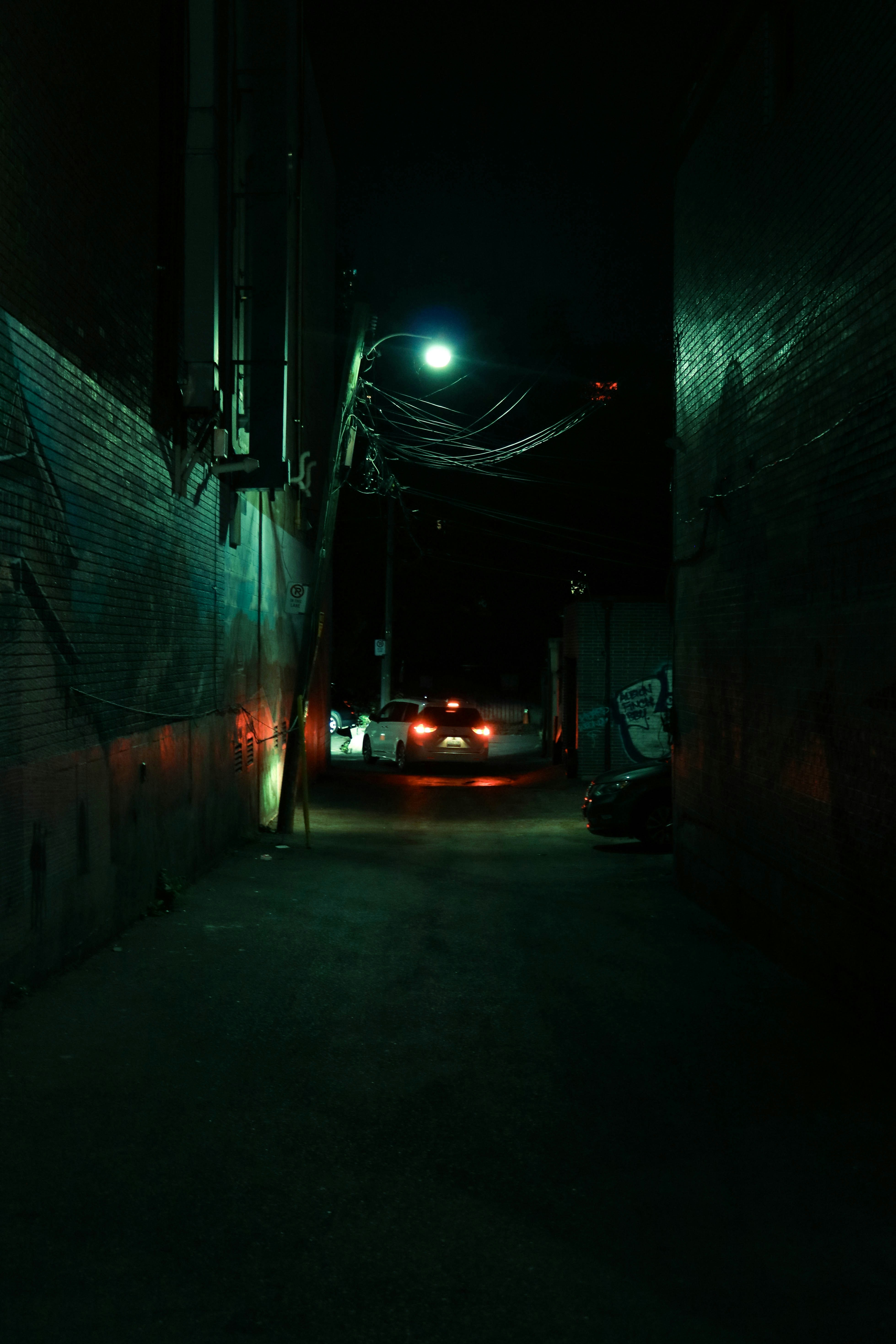 Dimly lit alleyway illuminated by a distant car's headlights, showcasing urban textures and shadows. The scene captures the essence of nighttime city life.