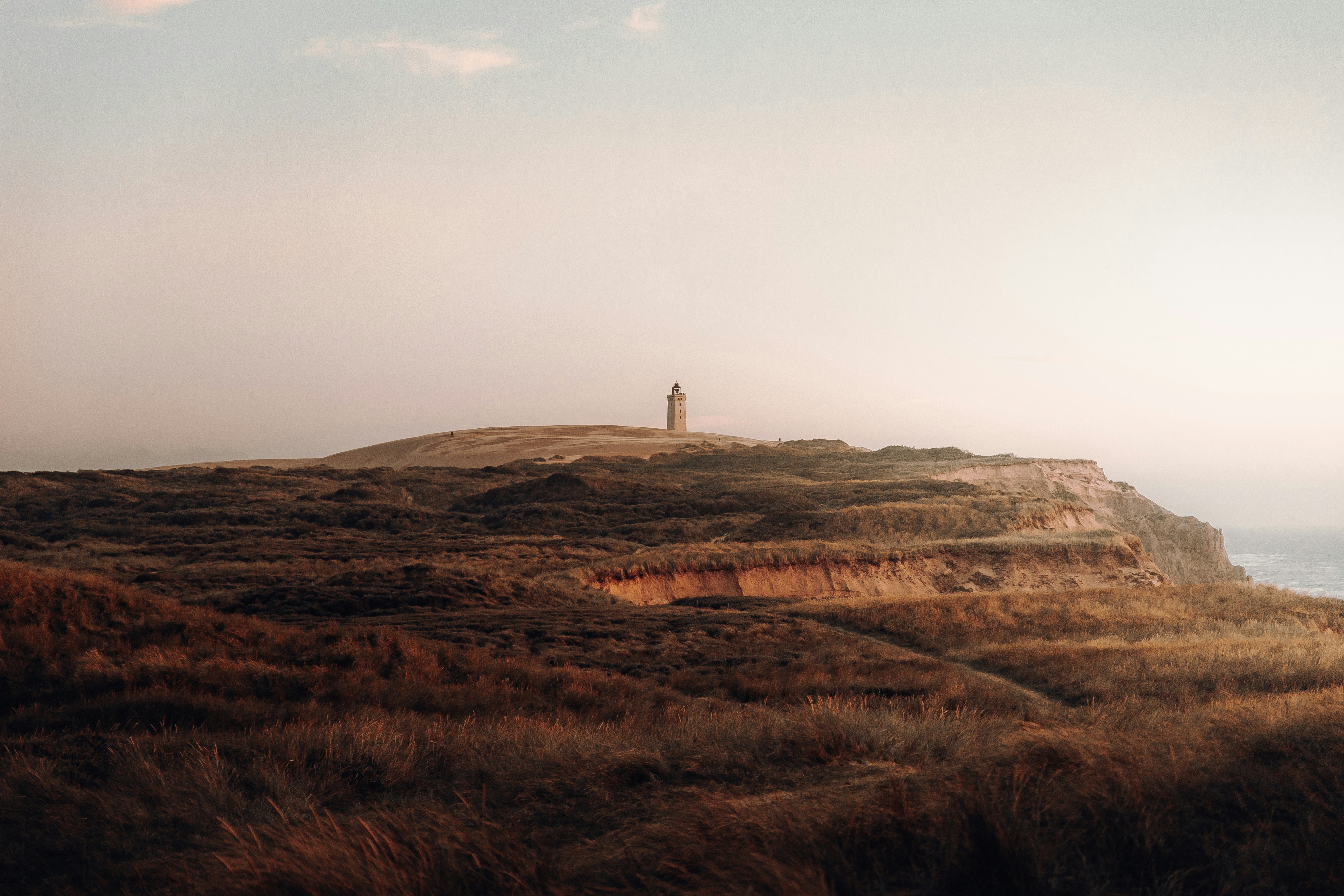 Lighthouse perched on a grassy cliff, surrounded by rolling hills and a serene coastal landscape at sunset.