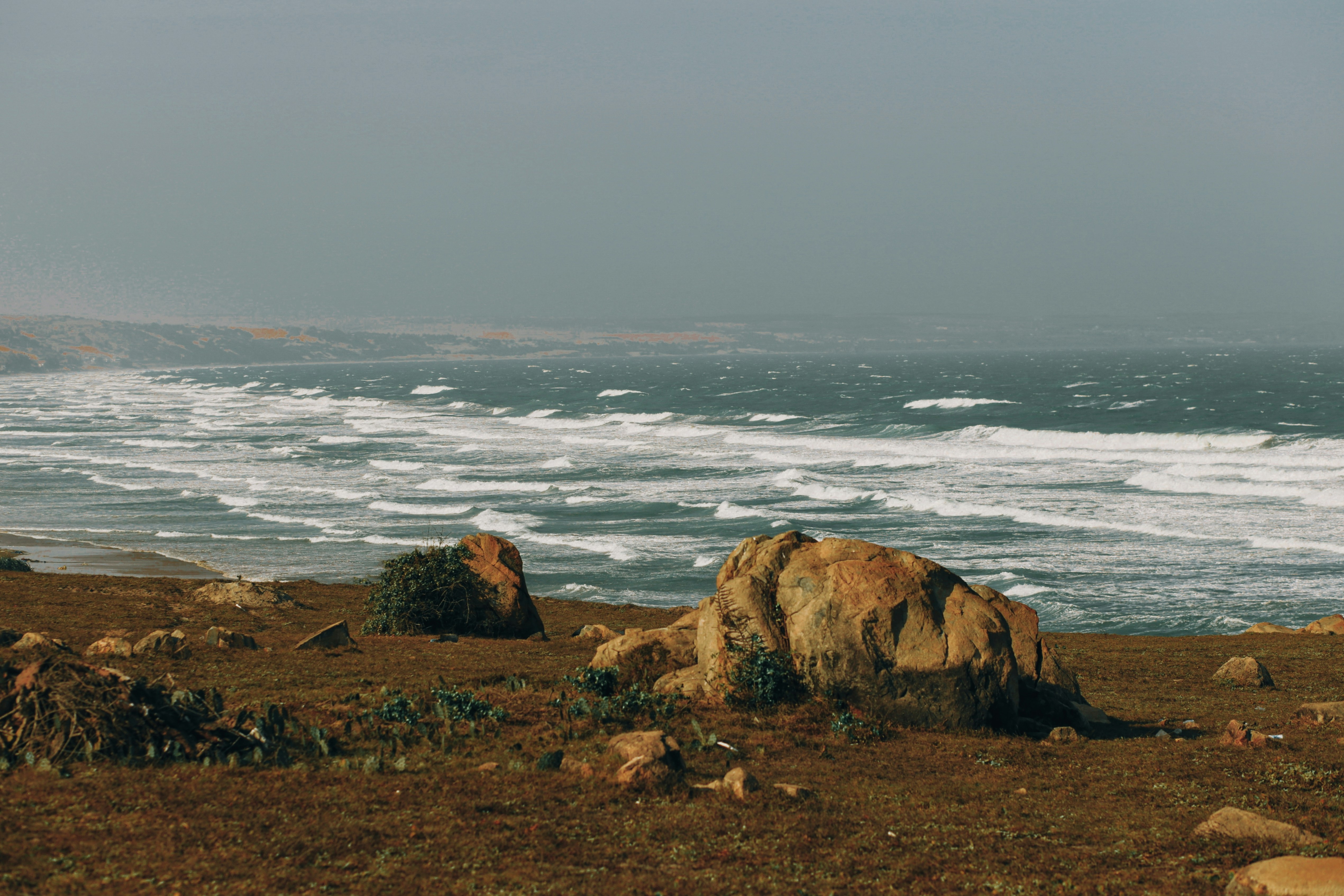 Waves crash against a rugged shoreline, with large boulders dotting the landscape under a moody sky.
