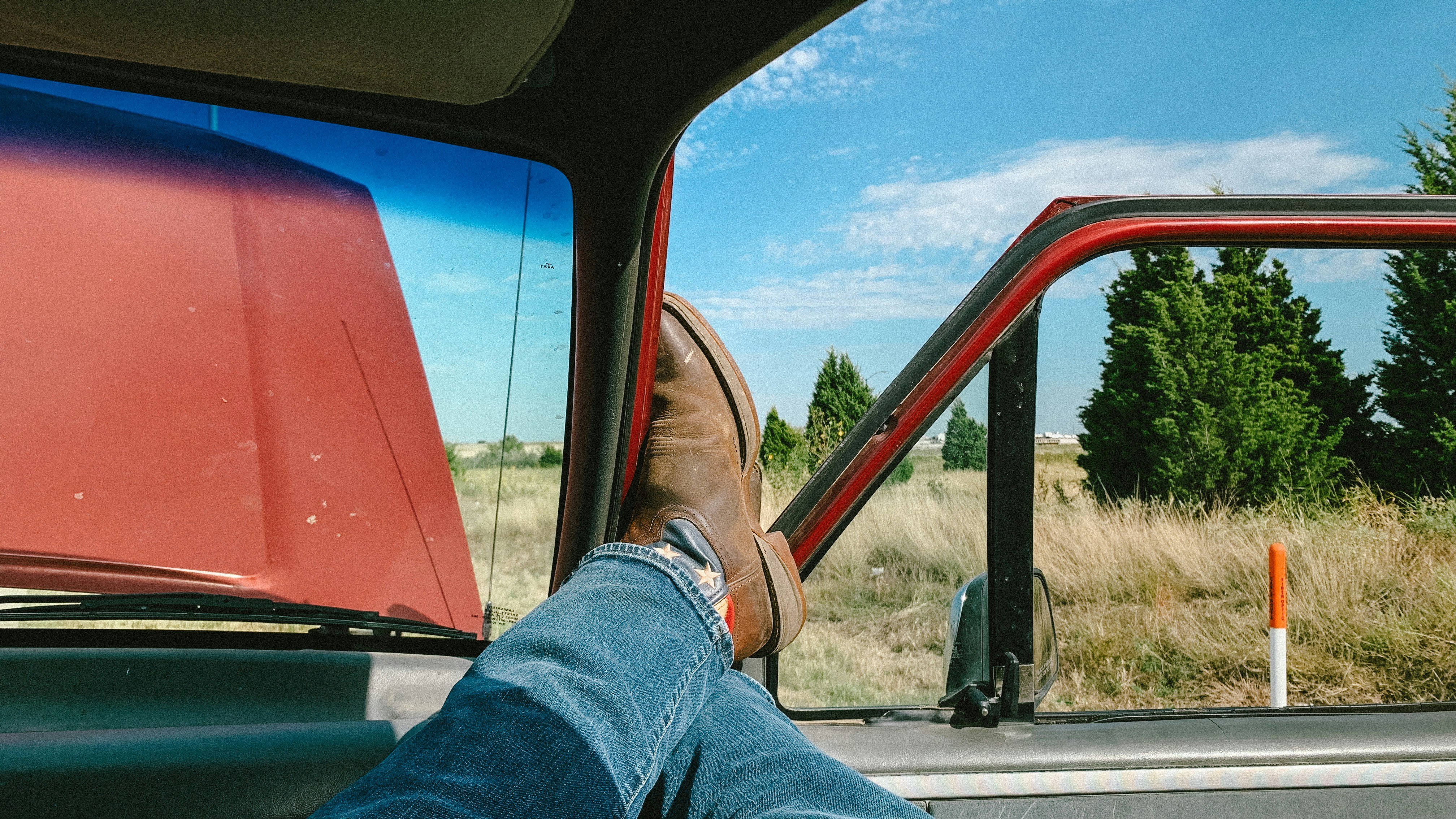 Foot resting on the dashboard of a truck with the hood raised, revealing a sunny landscape beyond. The scene captures a blend of leisure and automotive work.