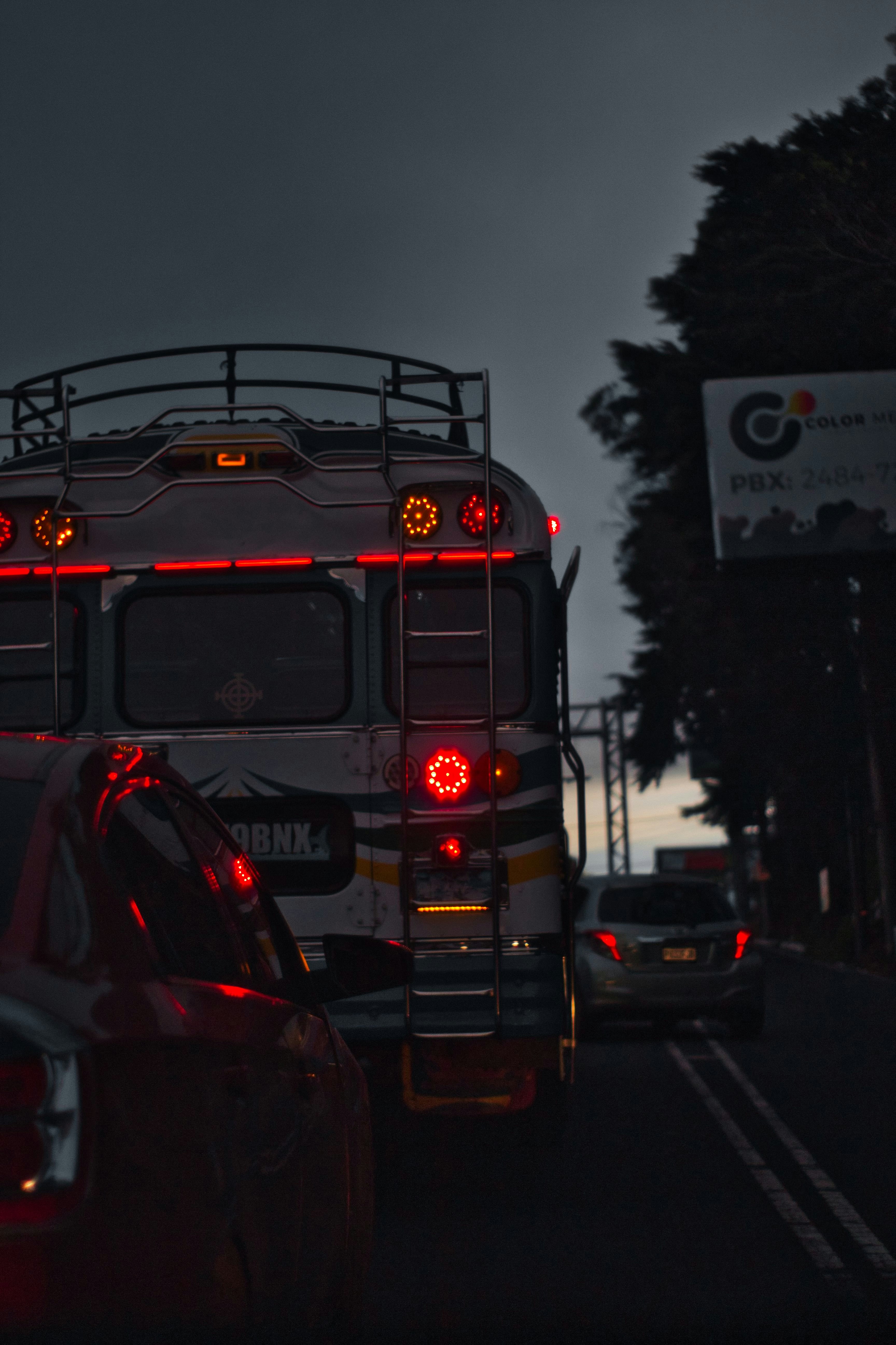 A colorful bus with illuminated lights is seen from behind, caught in evening traffic, surrounded by other vehicles. The scene captures the essence of urban commuting.
