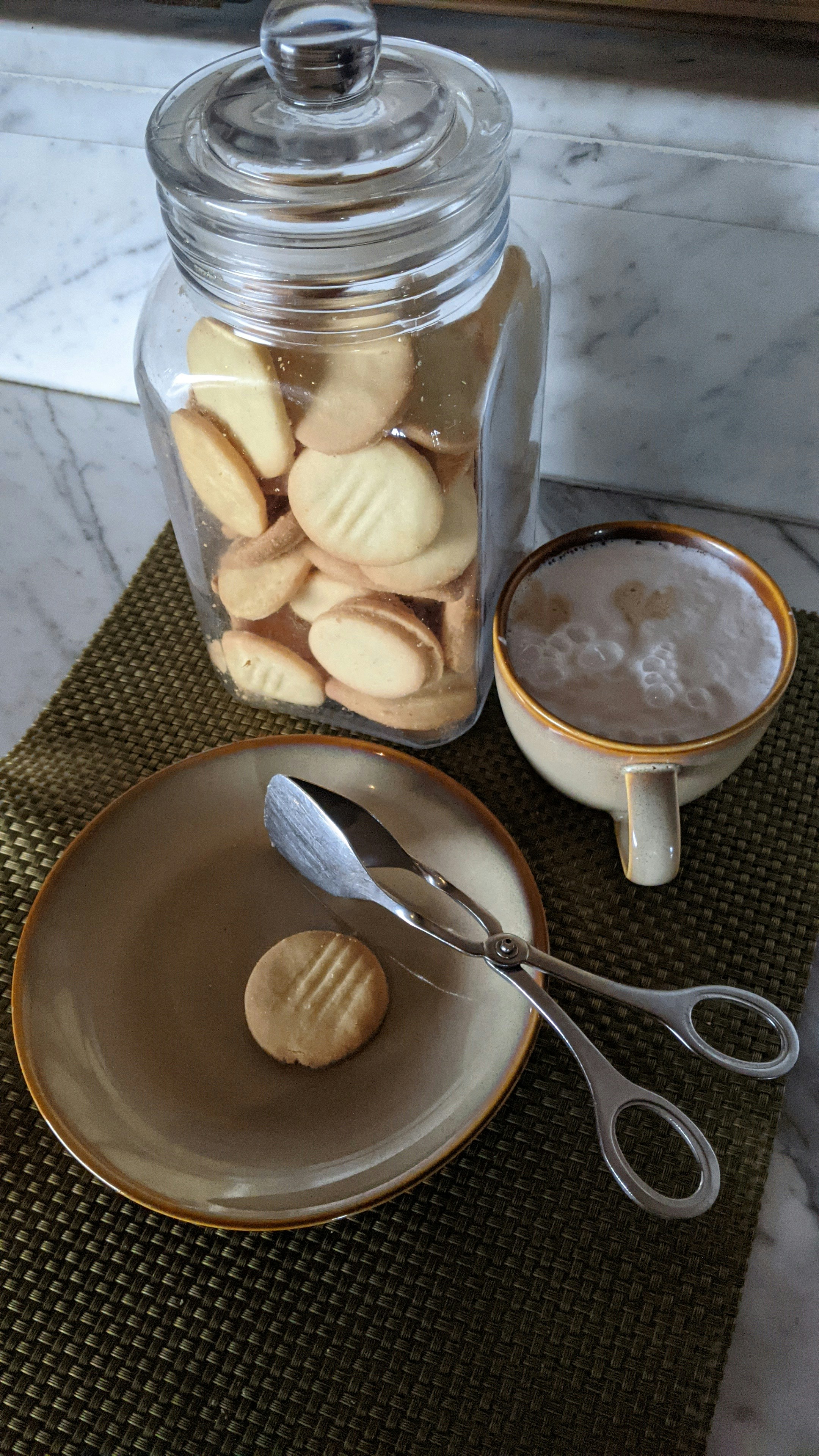 A jar filled with cookies sits beside a cup of frothy coffee on a textured mat, with a single cookie on a plate and a pair of scissors nearby.