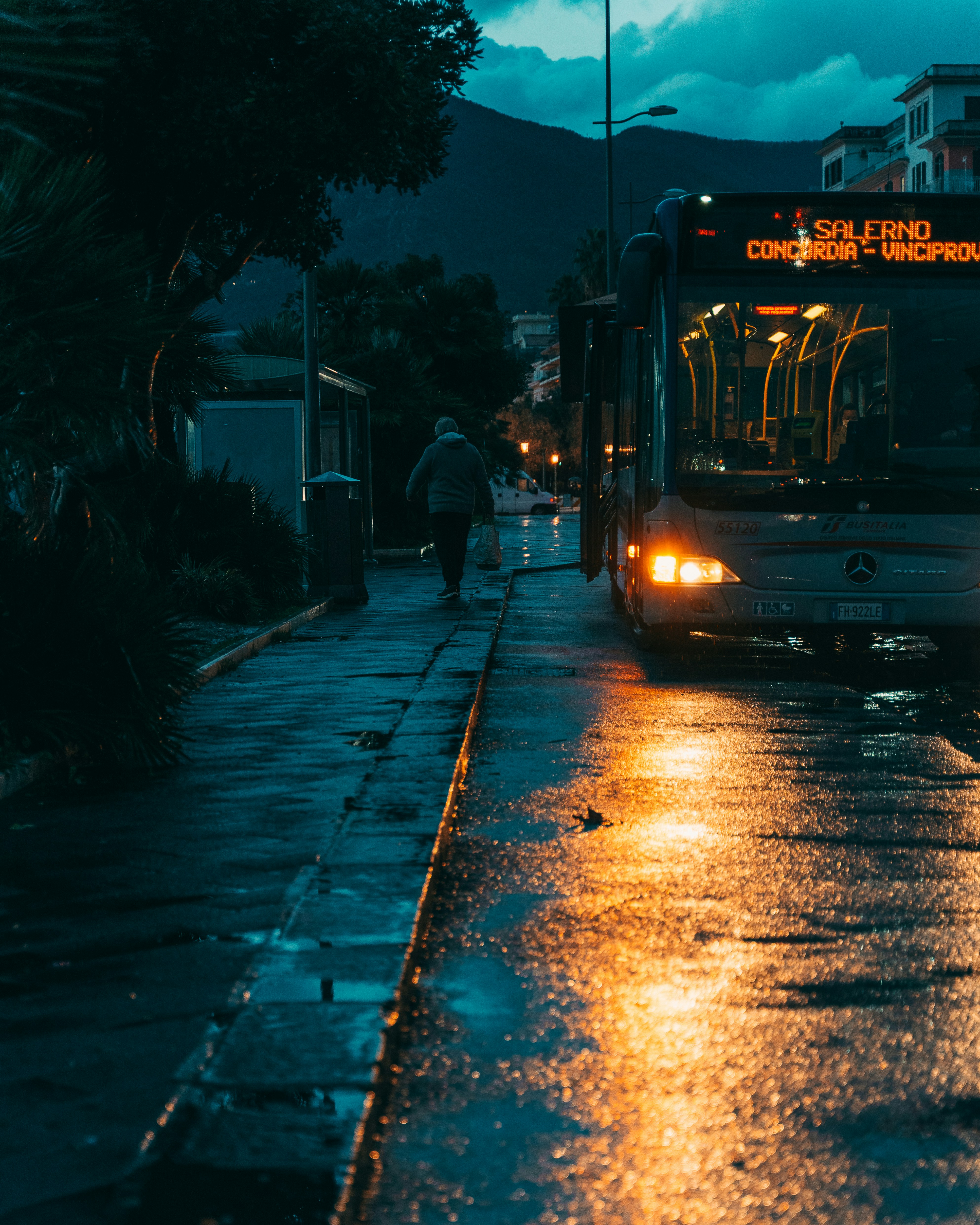 A city bus illuminated by streetlights navigates a rain-drenched sidewalk as a lone pedestrian walks by. The wet pavement reflects the warm glow of the bus's lights.