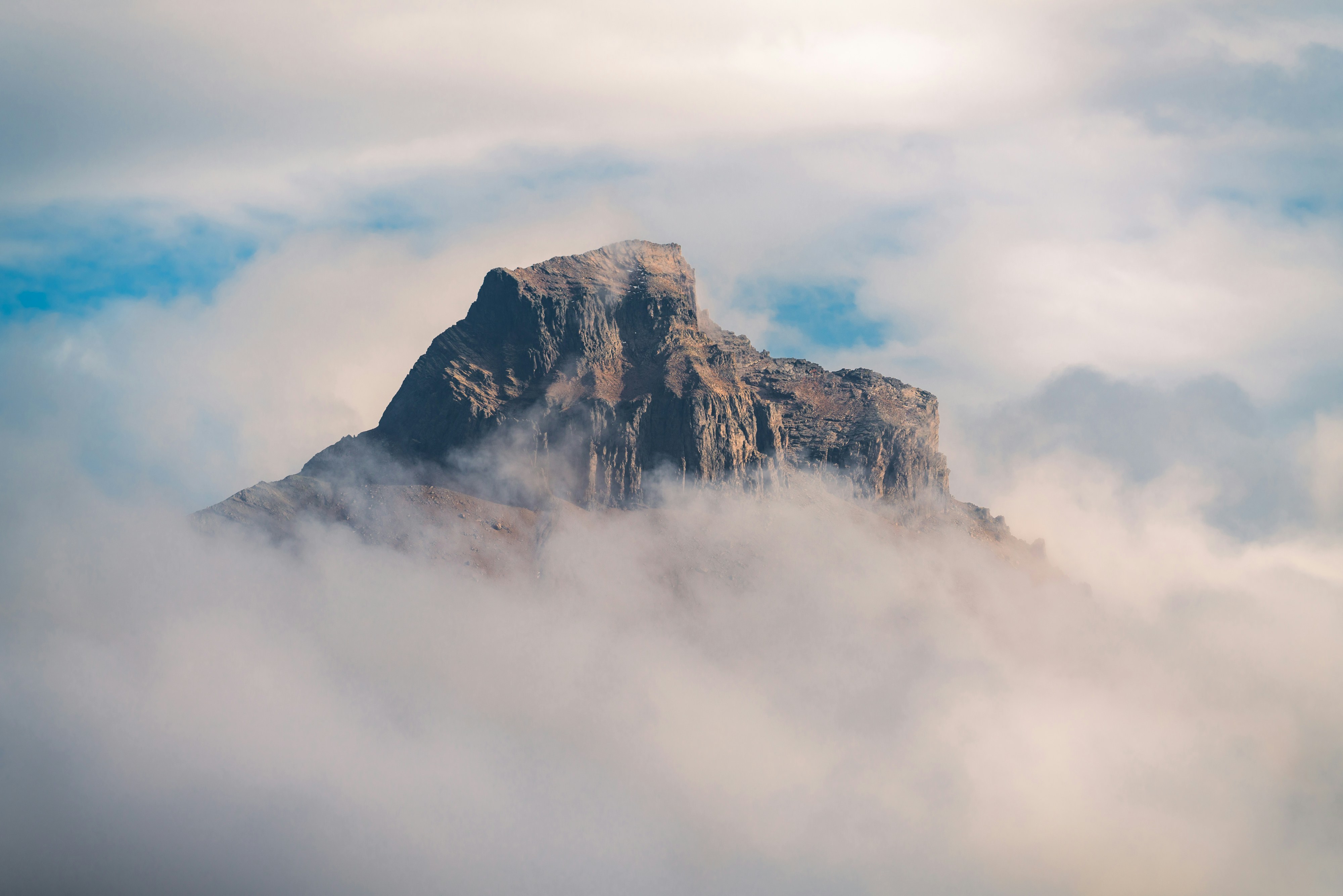 Majestic mountain peak emerging from a sea of clouds, surrounded by a soft blue sky. The rugged texture of the rock contrasts with the ethereal mist.