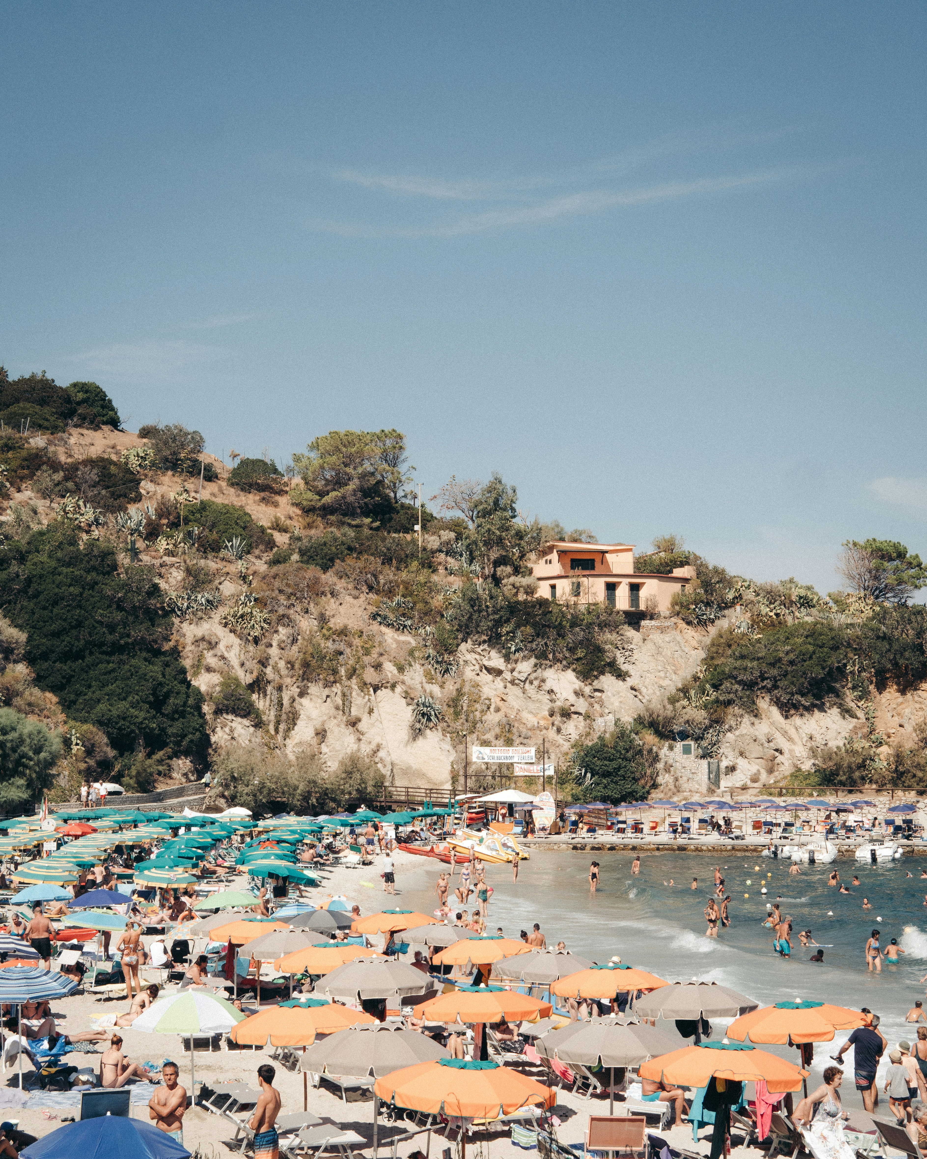 Vibrant beach scene with colorful umbrellas and sunbathers enjoying the sun, framed by a rocky cliff and lush greenery.