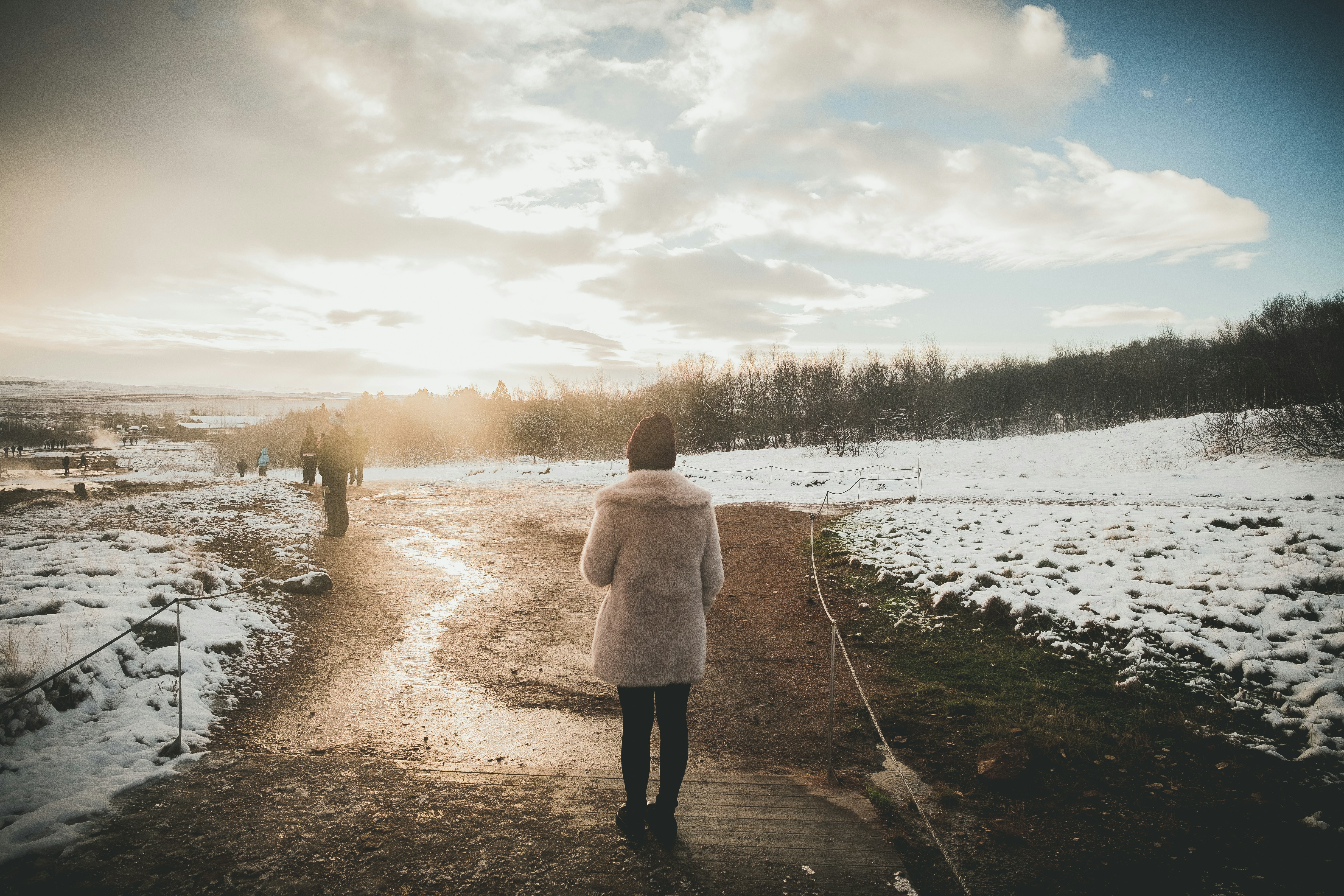A solitary figure walks along a snow-covered trail under a cloudy sky, with the sun setting in the distance. The scene captures the serene beauty of winter's embrace.