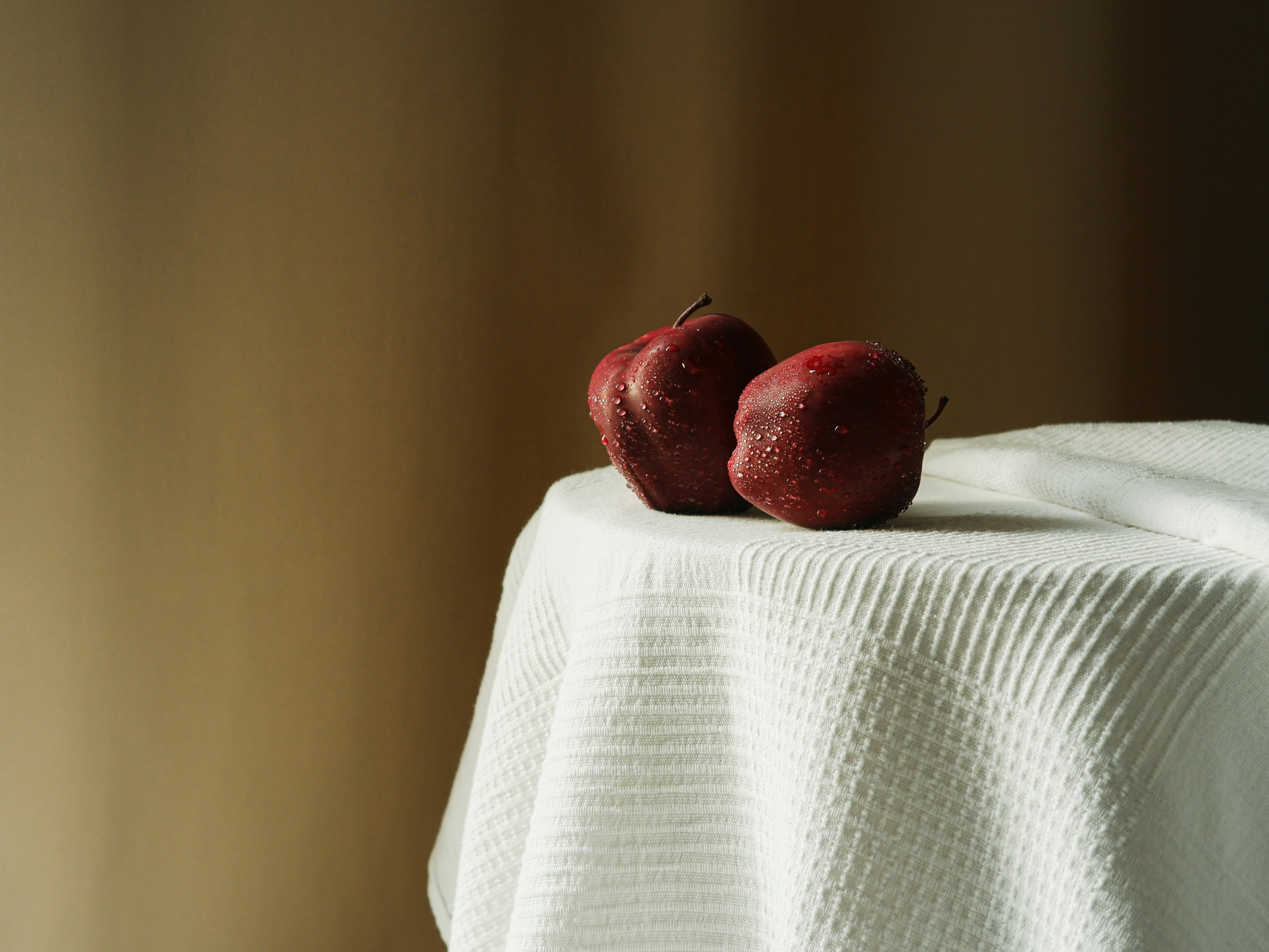 Two red cherries rest on a white textured cloth, forming a simple still life. The shallow depth of field keeps the fruit in sharp focus while the background remains softly blurred.