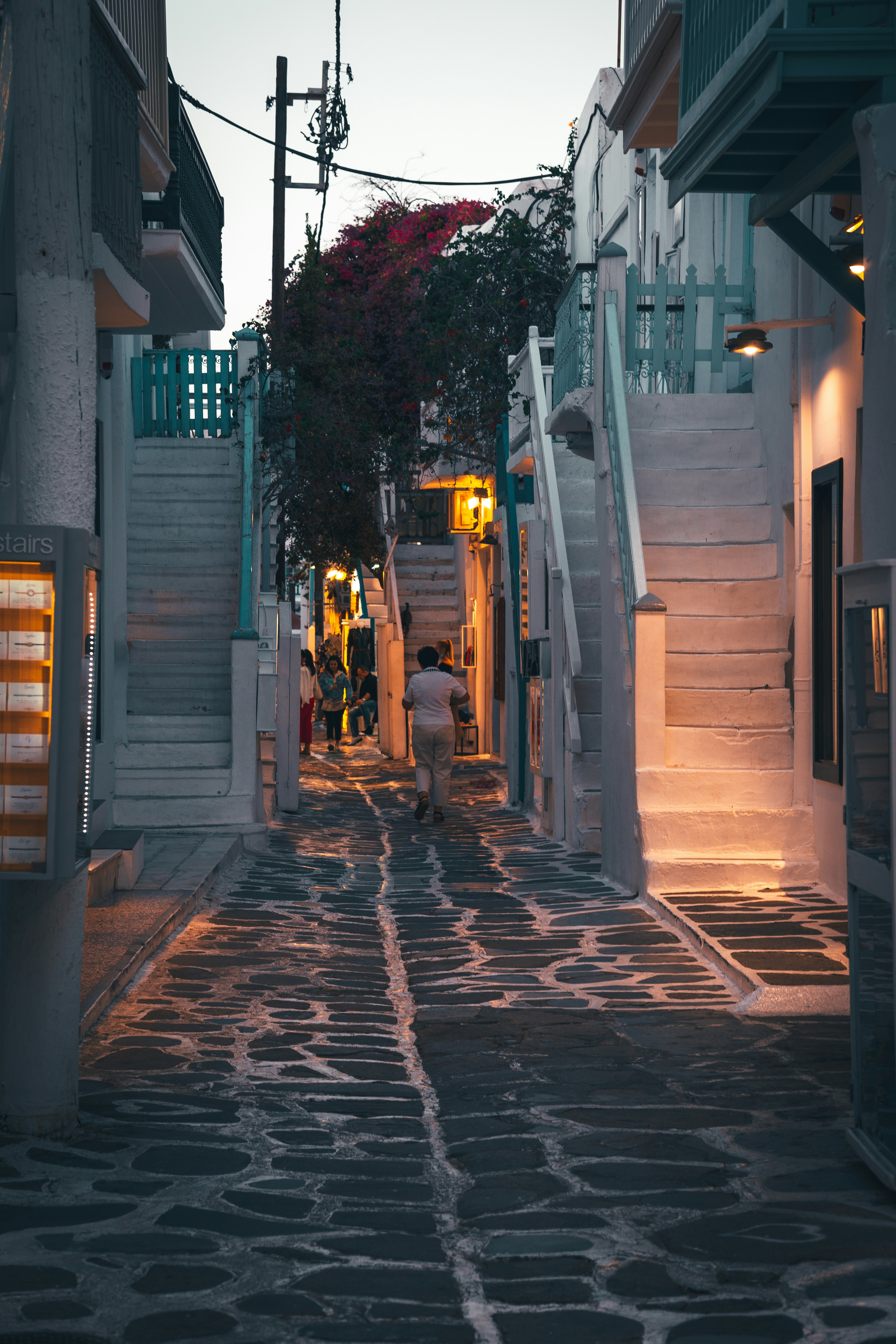 A quaint alleyway illuminated by warm streetlights, lined with whitewashed buildings and vibrant bougainvillea. The cobblestone path invites evening wanderers.