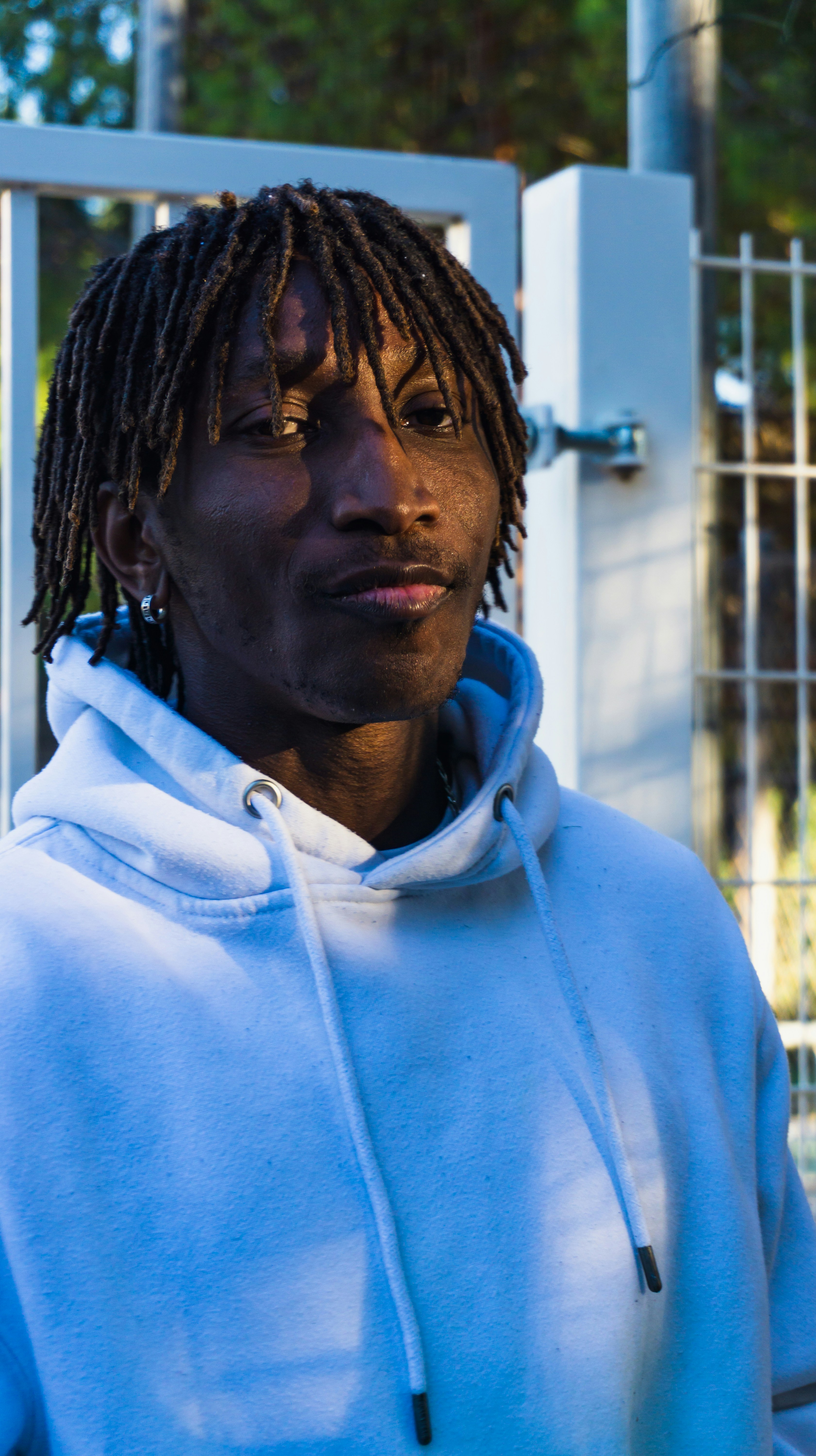 A young man with braided hair gazes thoughtfully, framed by a backdrop of greenery and a fence. The moment captures a blend of contemplation and connection to the environment.