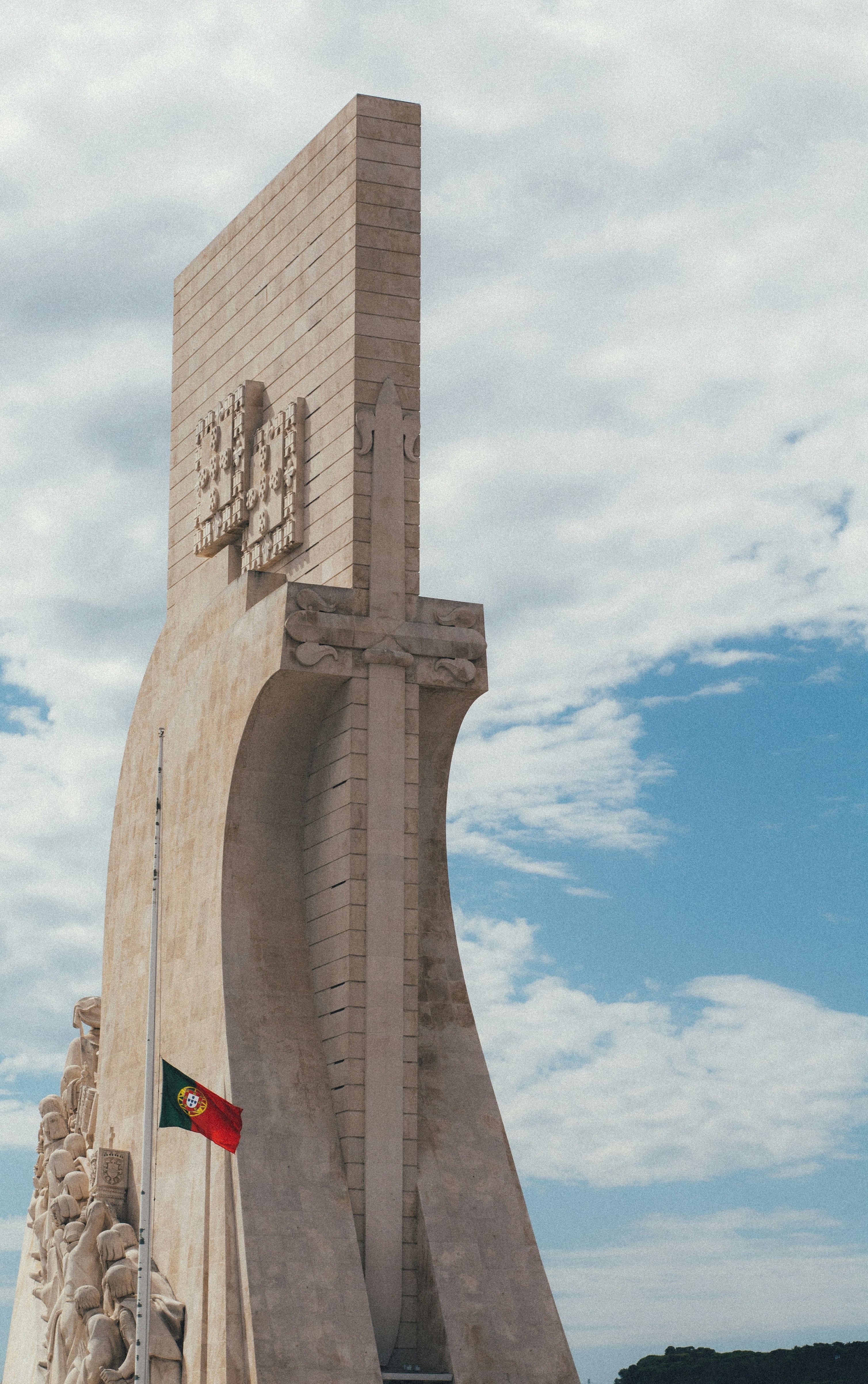 A towering stone monument featuring intricate carvings and the Portuguese flag, set against a backdrop of a cloudy sky.