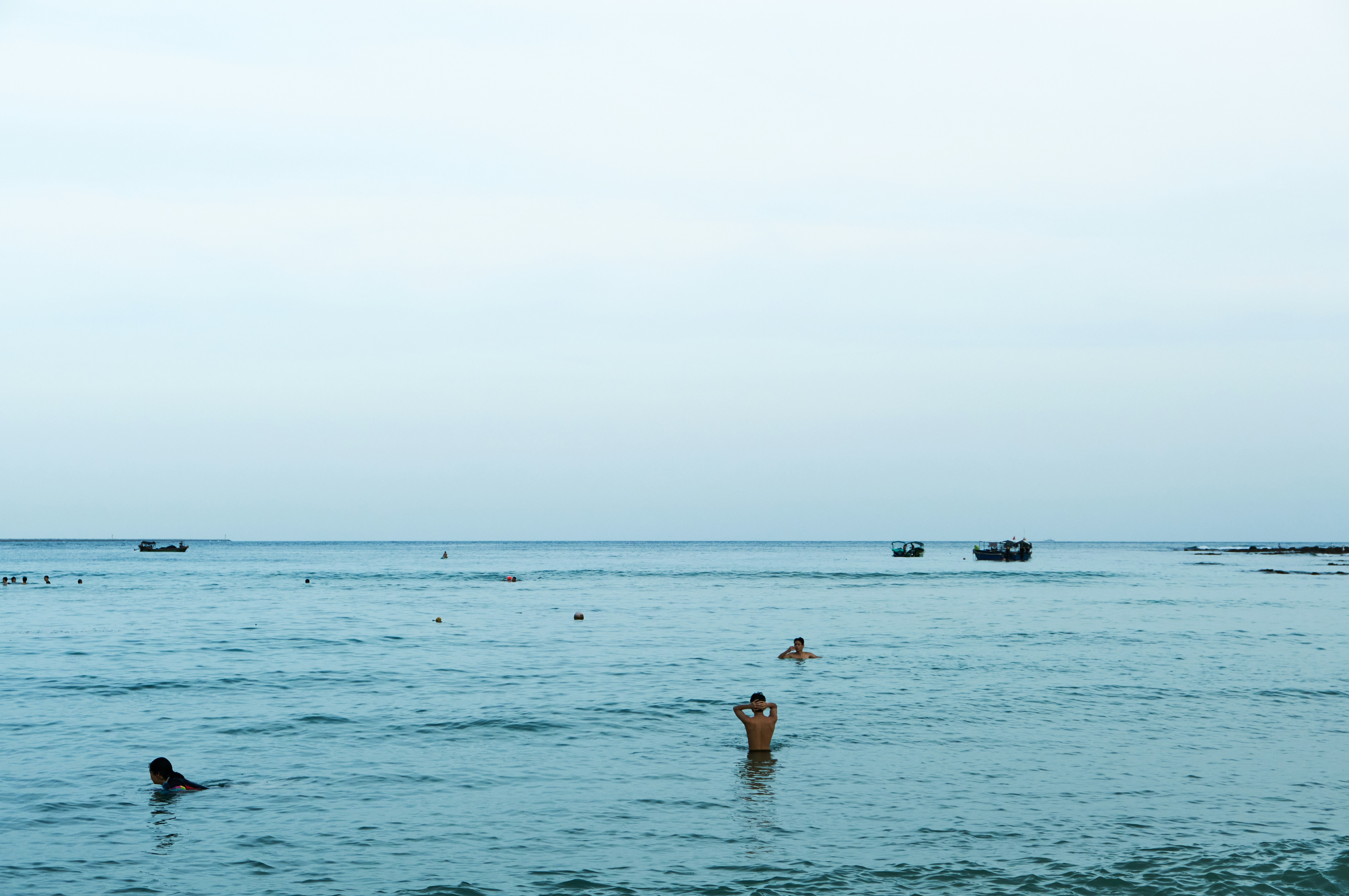 Swimmers enjoying a calm sea under a soft blue sky, with distant boats dotting the horizon.