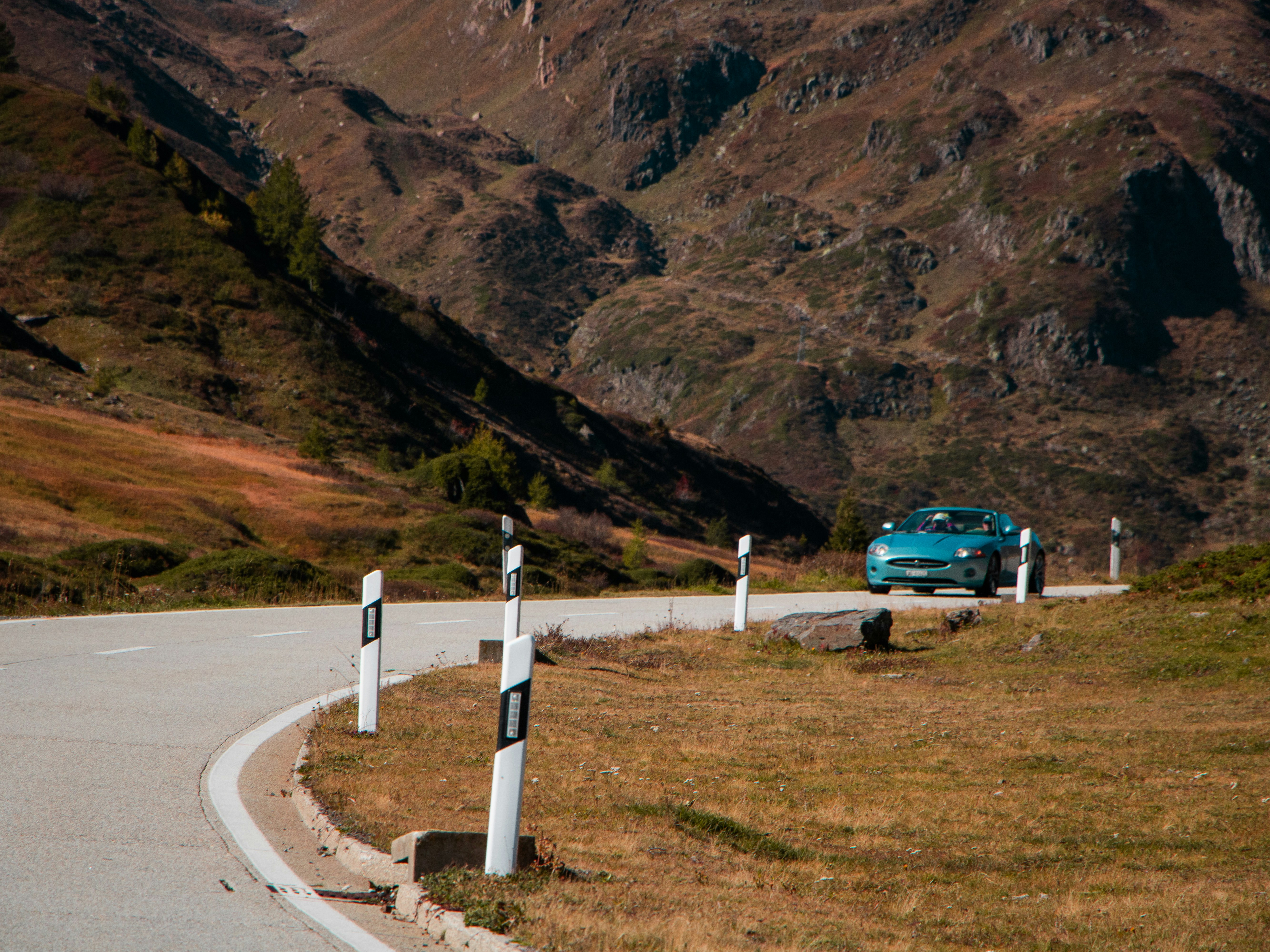 A turquoise sports car navigating a winding mountain road surrounded by rugged terrain and autumn foliage.