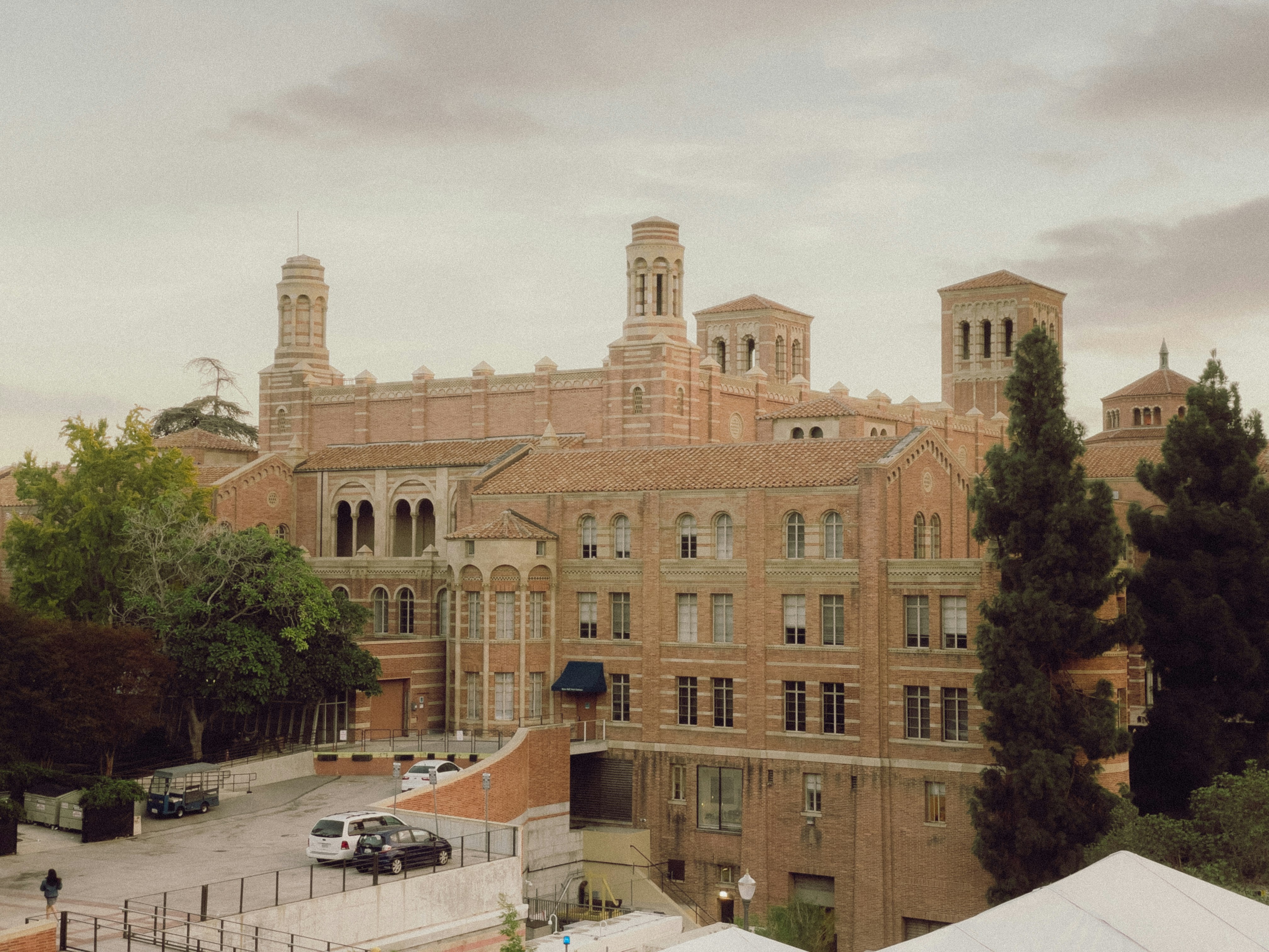 Historic brick building with intricate architectural details surrounded by lush greenery and vehicles parked below.