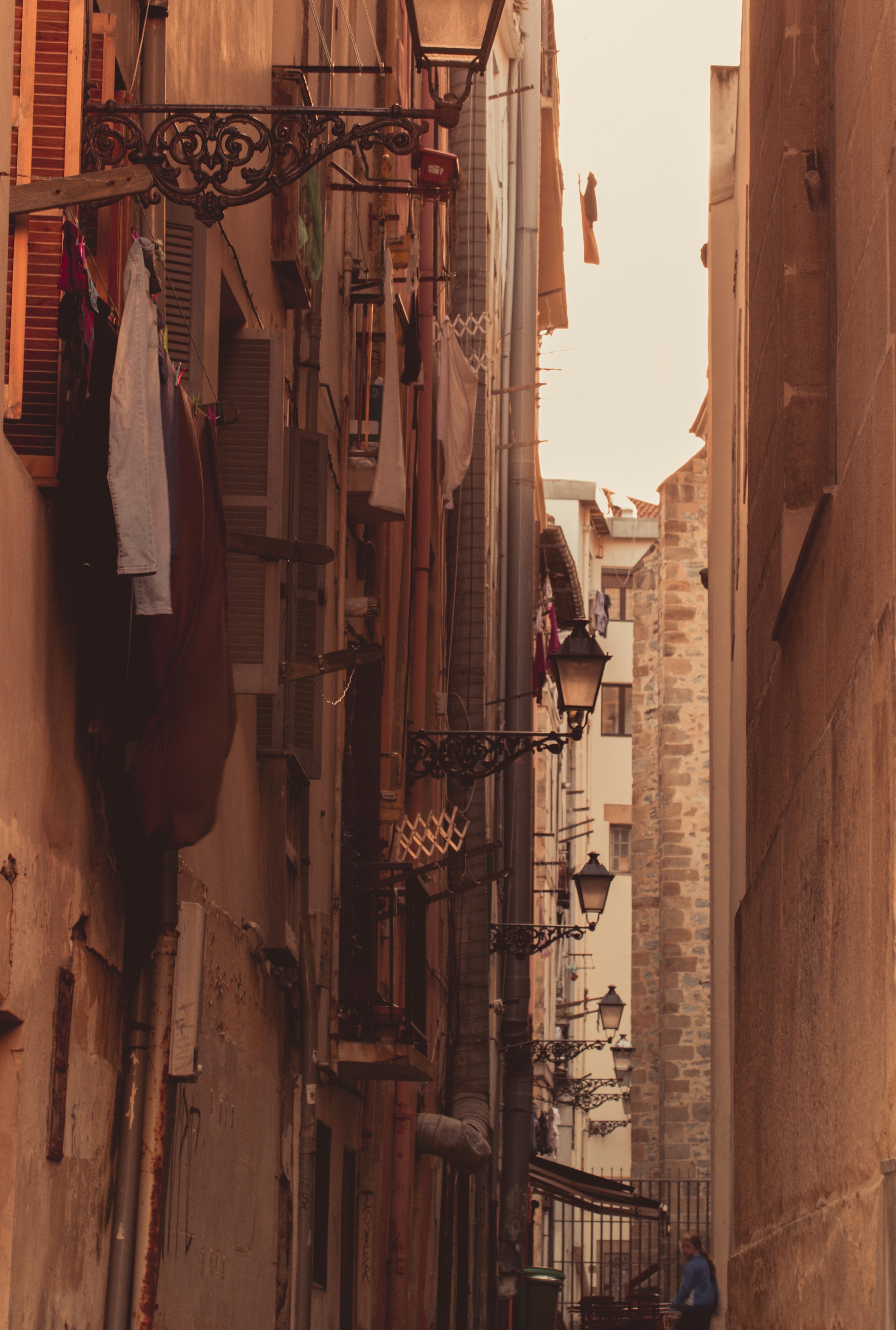 Clothes hang between weathered walls in a narrow alley, illuminated by soft evening light. Vintage street lamps add character to the scene.