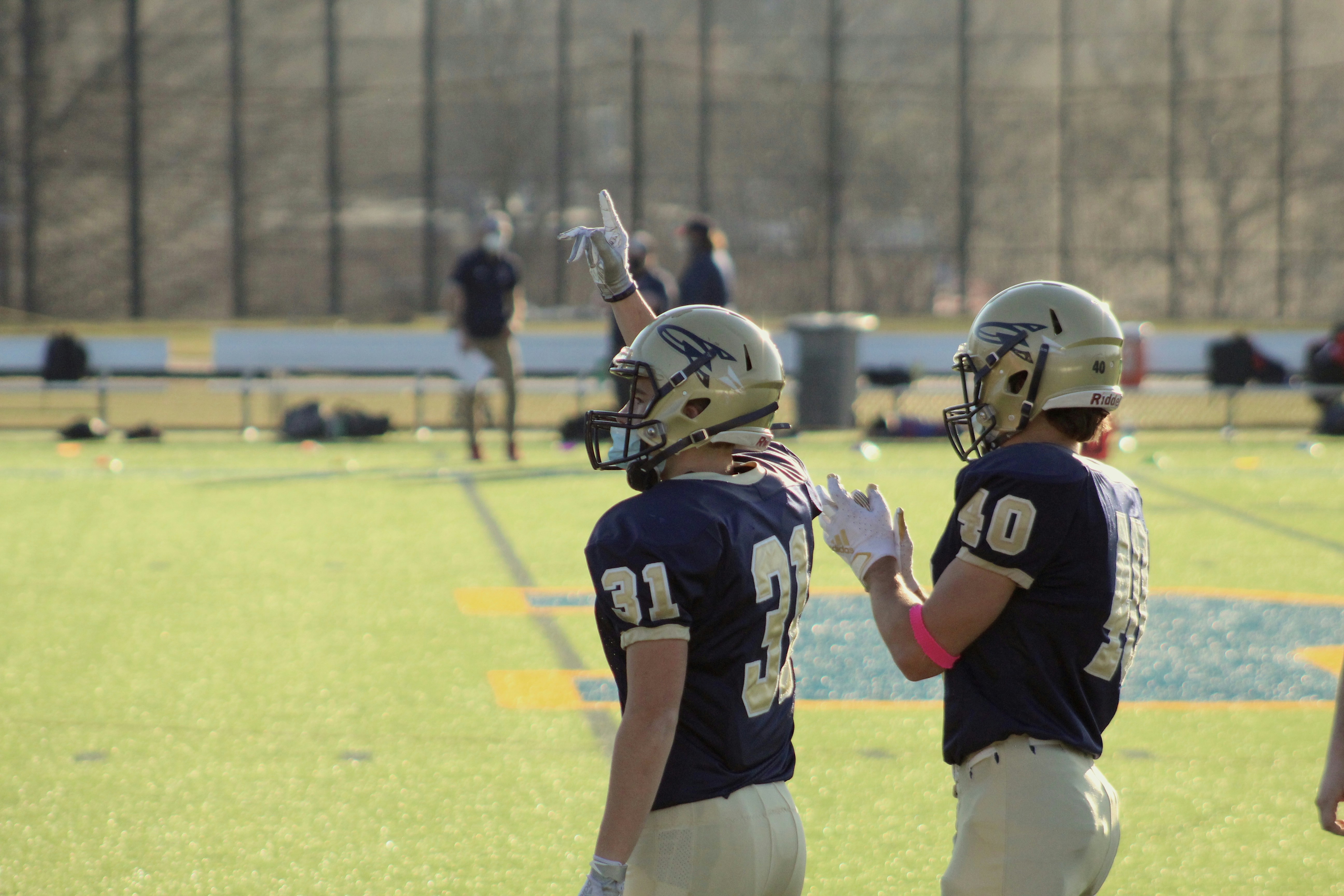 Two football players in blue uniforms stand on a grass field under soft sunlight.
