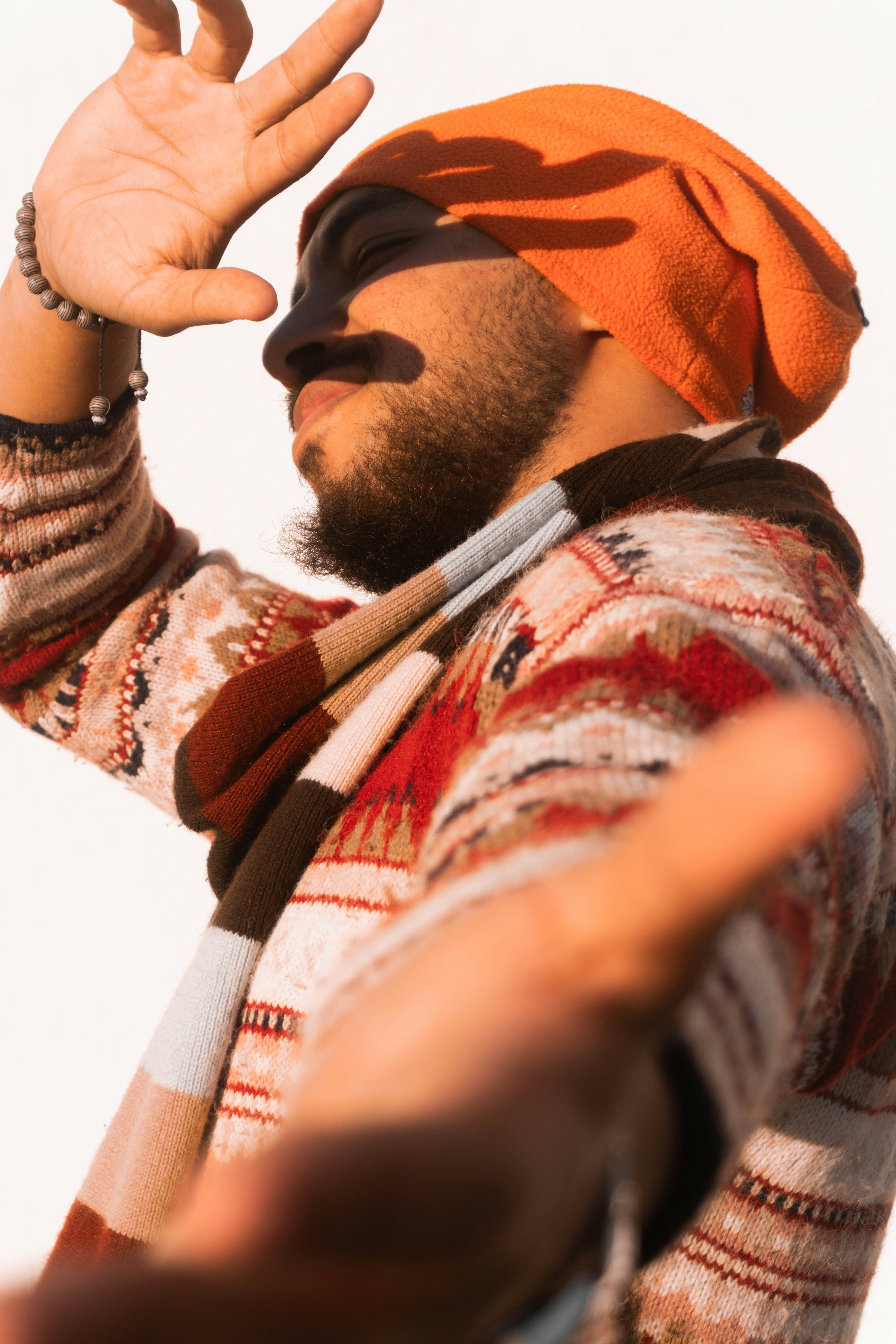 A man in a colorful sweater and orange beanie extends his hands outward, creating a sense of connection and warmth against a bright background.
