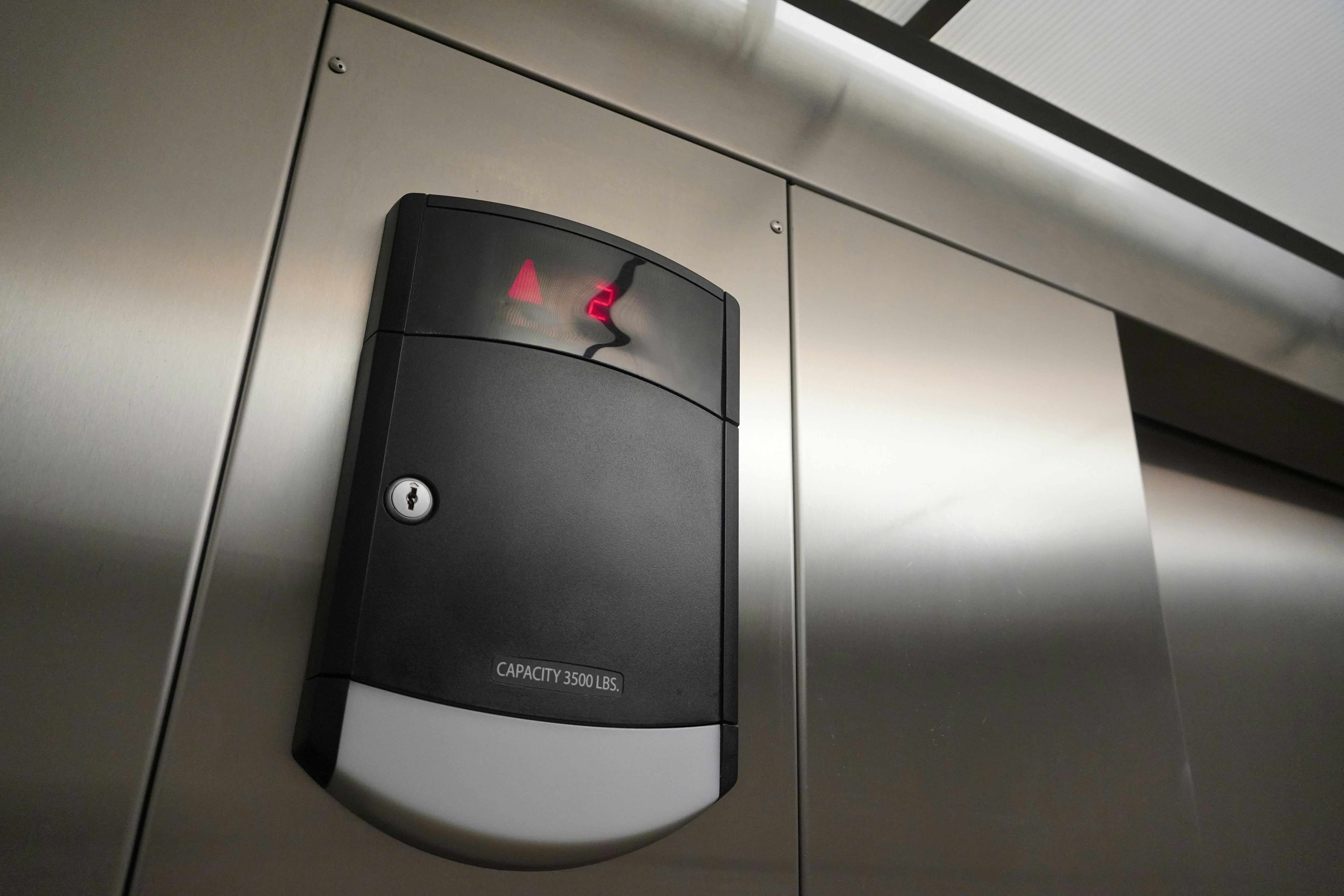 Elevator control panel displaying upward and downward arrows along with weight capacity information. The sleek metallic surface reflects the modern design of urban architecture.