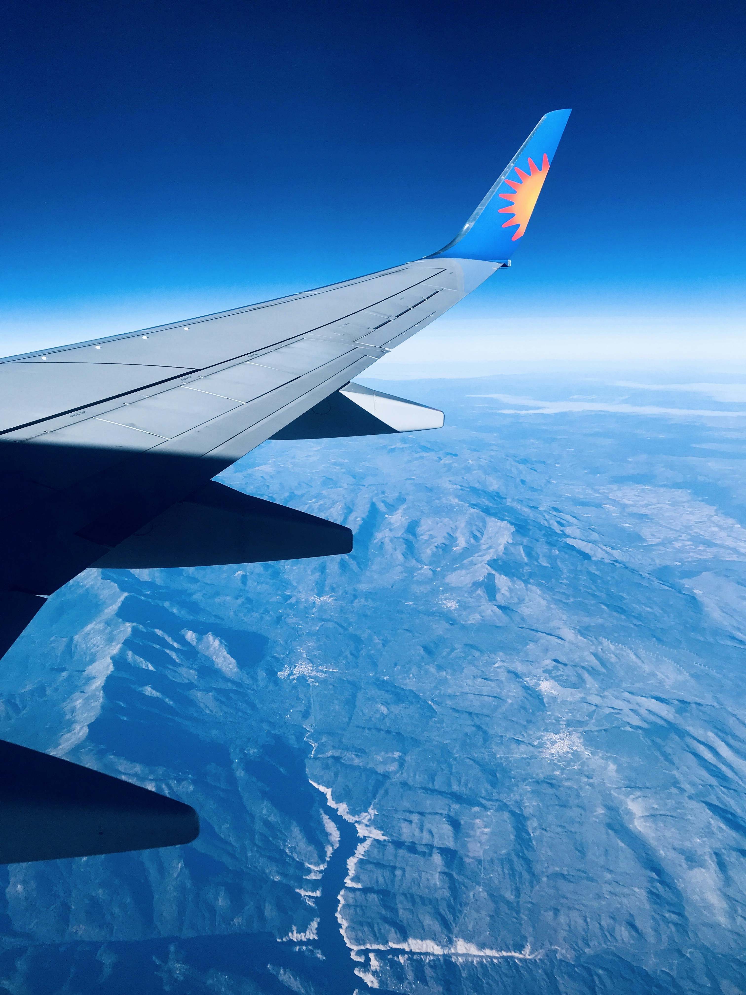 Airplane wing extending into a clear blue sky, showcasing distant mountain ranges and valleys below.