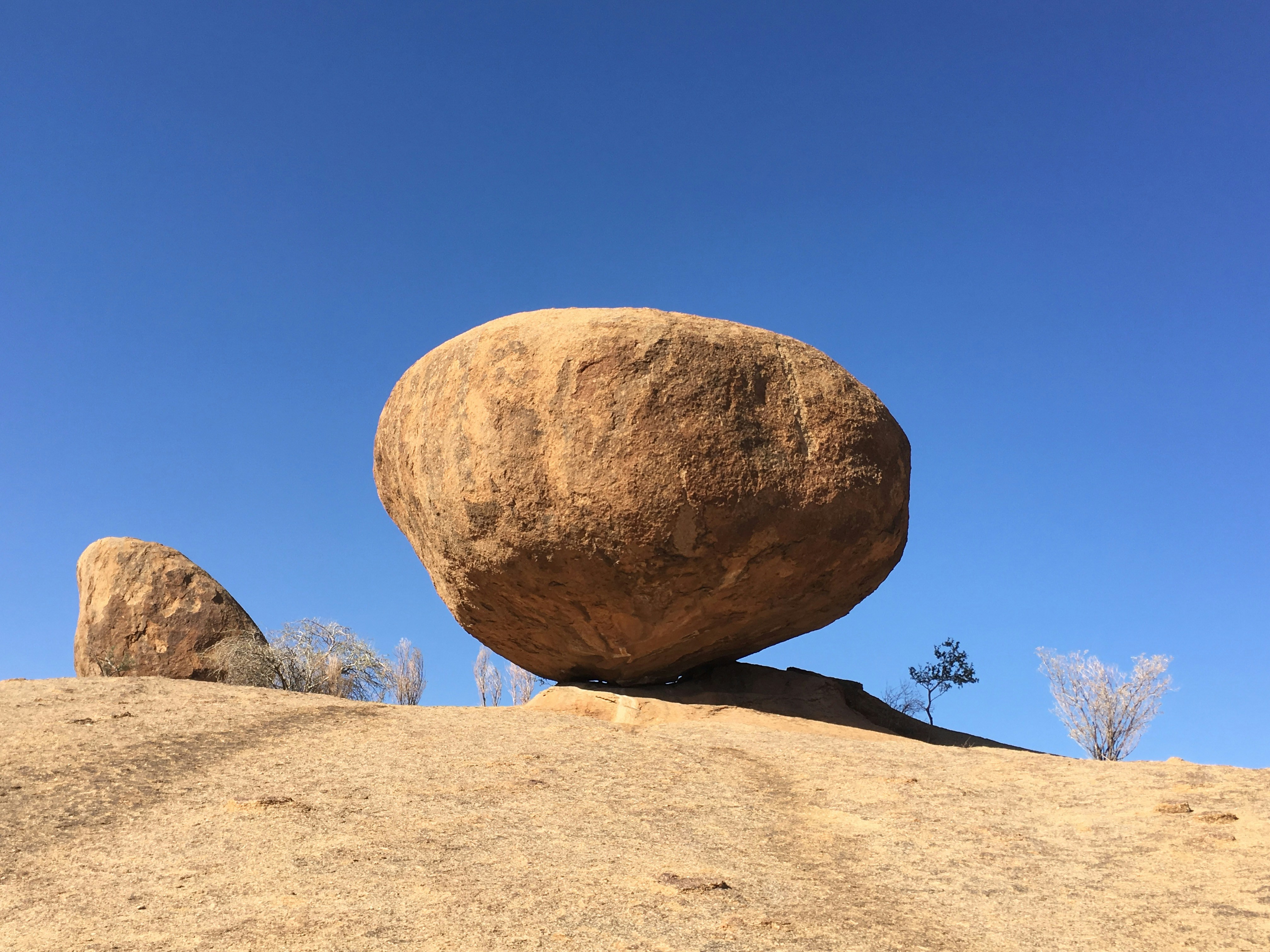 A large boulder precariously balanced on a smaller rock formation under a clear blue sky. The surrounding landscape features dry grass and sparse vegetation.