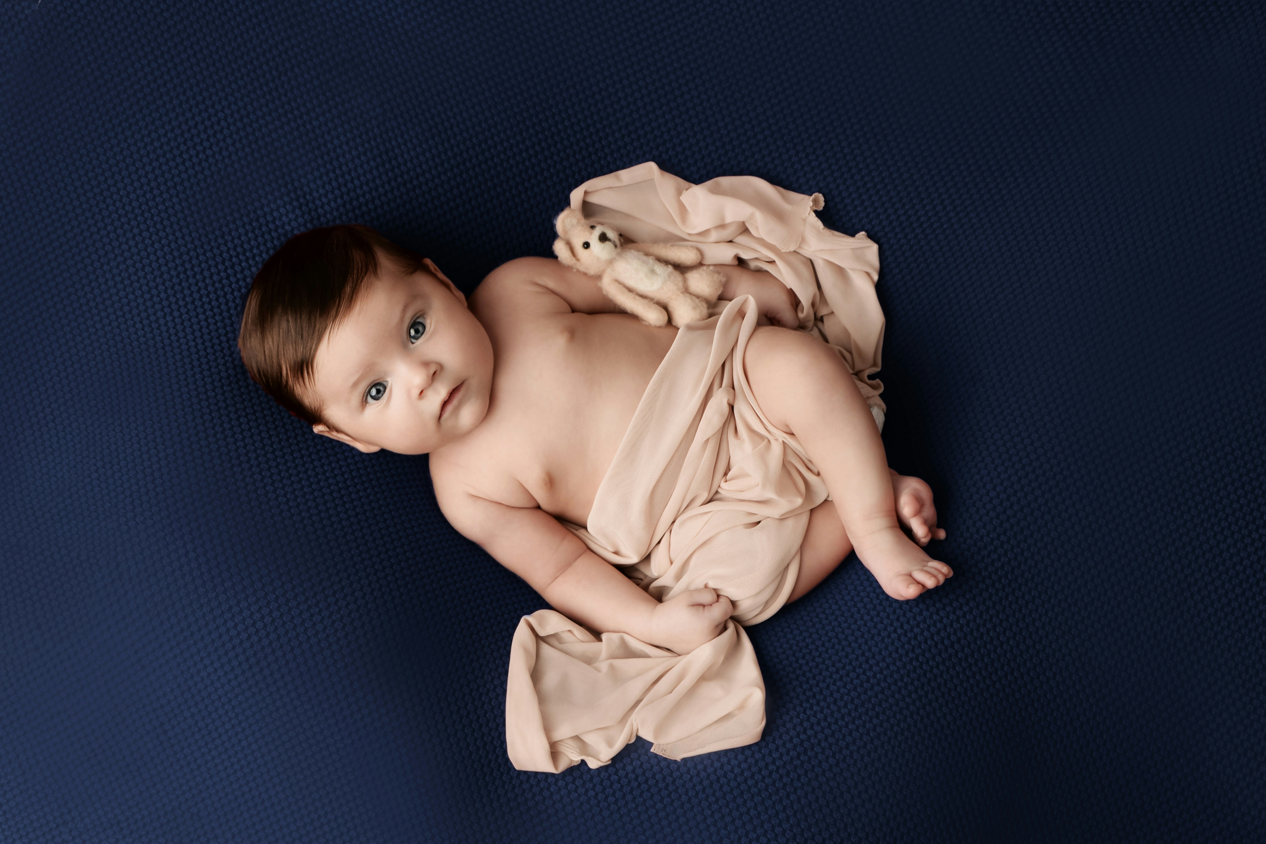 Newborn baby wrapped in soft fabric, holding a small teddy bear, set against a deep blue background.